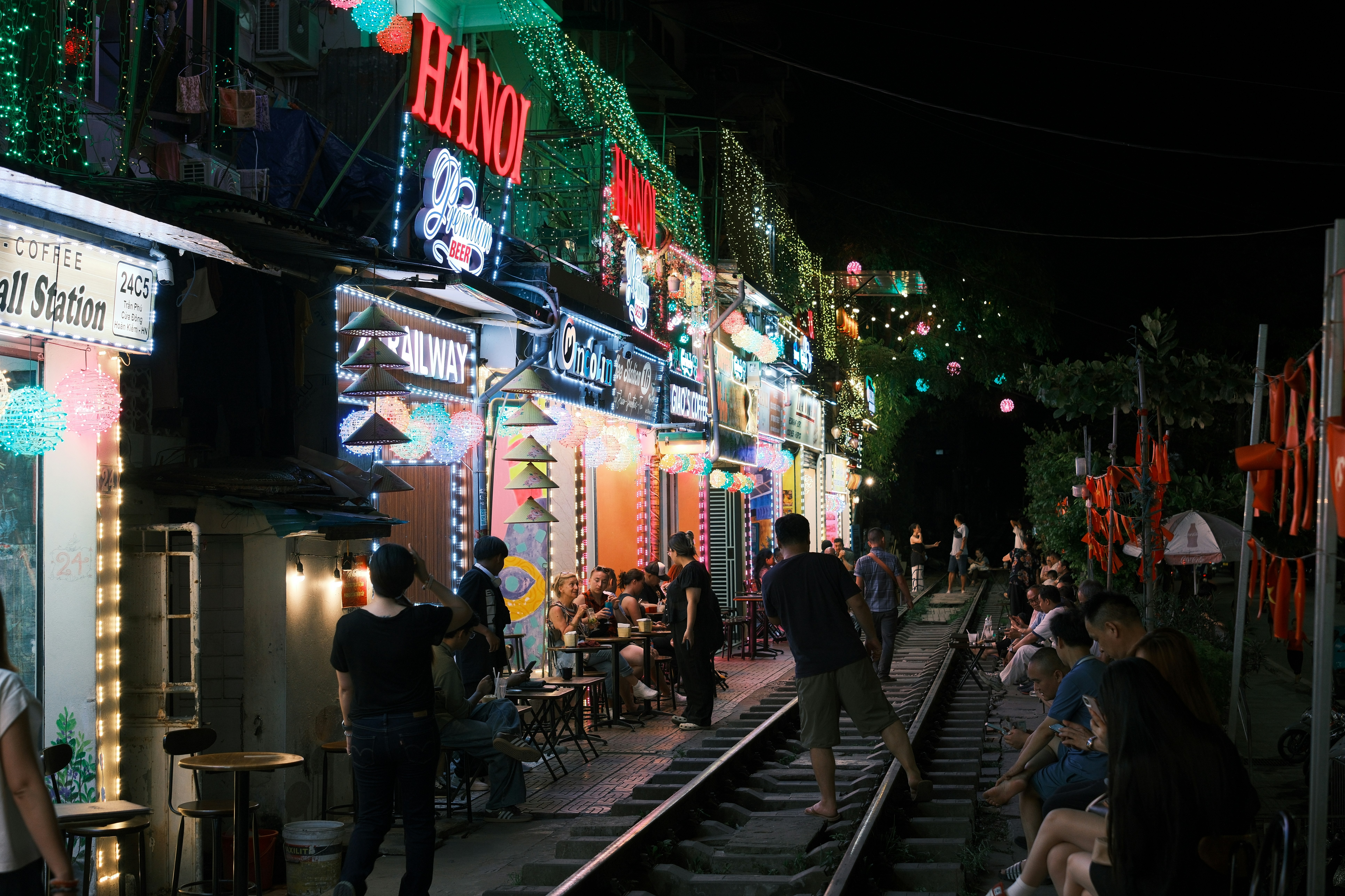 Vibrant street scene along a railway in Hanoi, illuminated by colorful neon lights and bustling with people enjoying the nightlife.