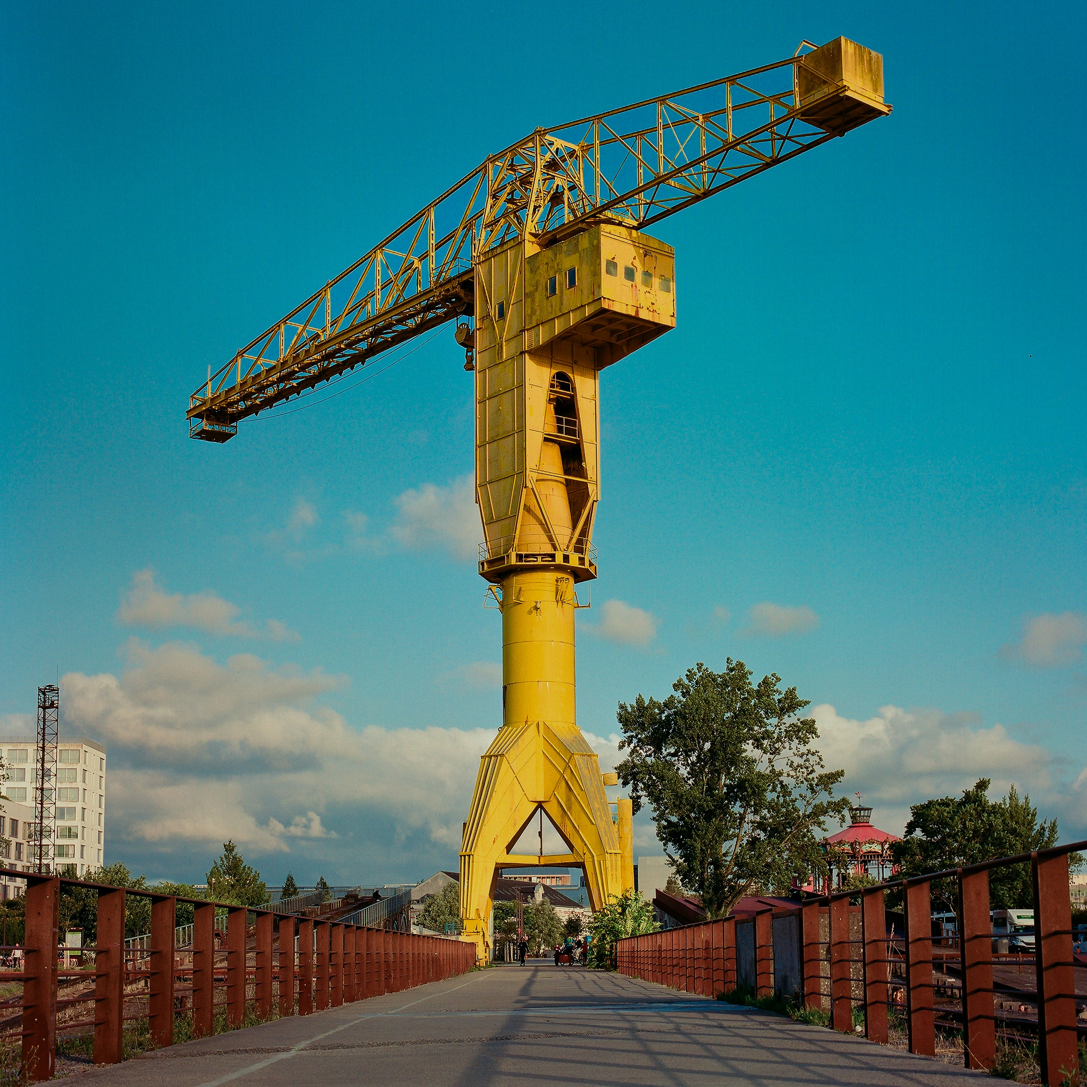 The iconic yellow Titan crane in Nantes, France, photographed on Kodak Portra 160 film with a Hasselblad 500 C/M. This striking industrial structure stands tall against a deep blue summer sky, echoing the city’s shipbuilding heritage. Shot in July 2025 from the pedestrian bridge that leads directly to its base, this composition highlights the crane’s architectural power and historical significance. | Yellow crane stands tall under a blue sky.