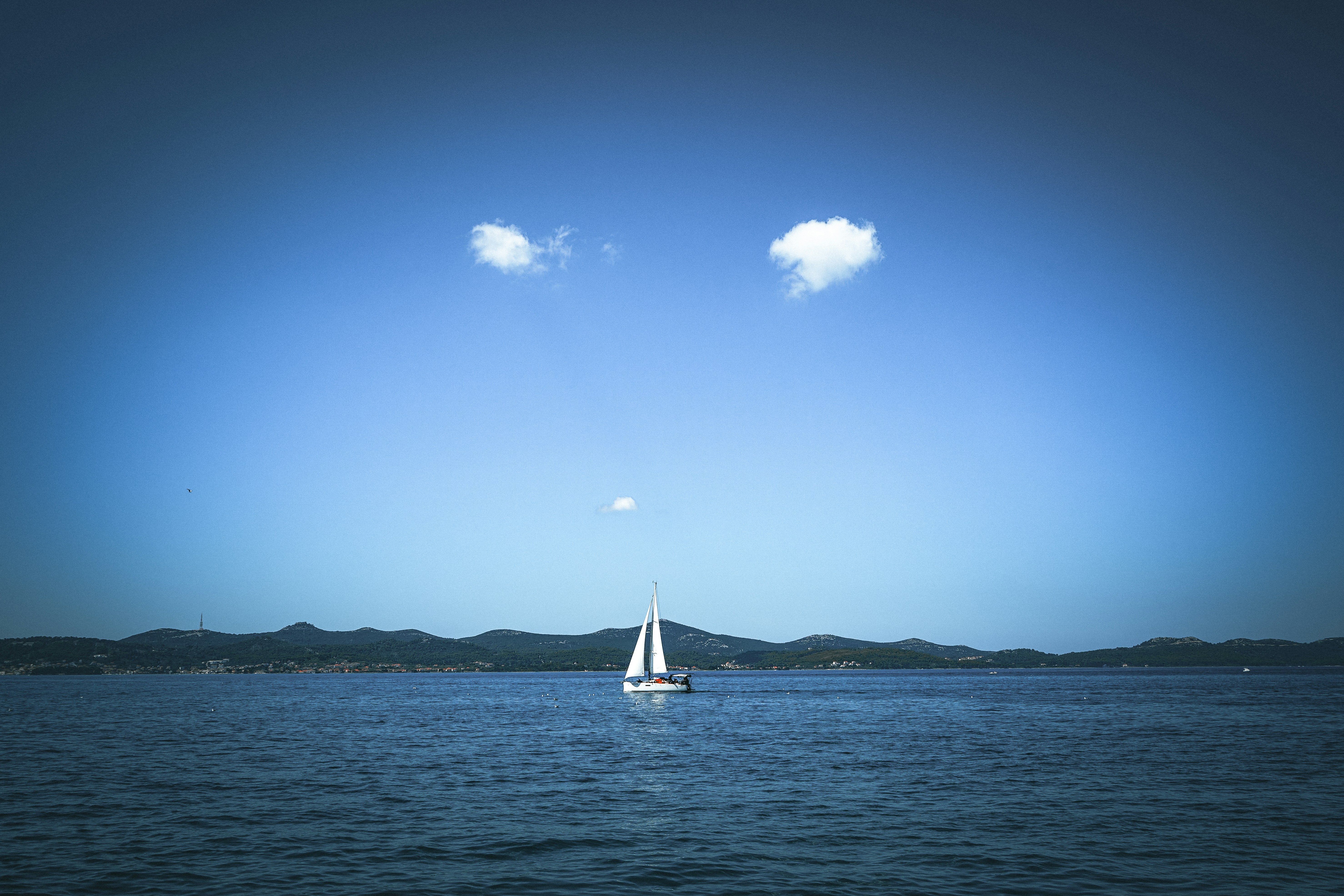Sailboat gliding across calm waters under a clear blue sky with distant hills on the horizon.