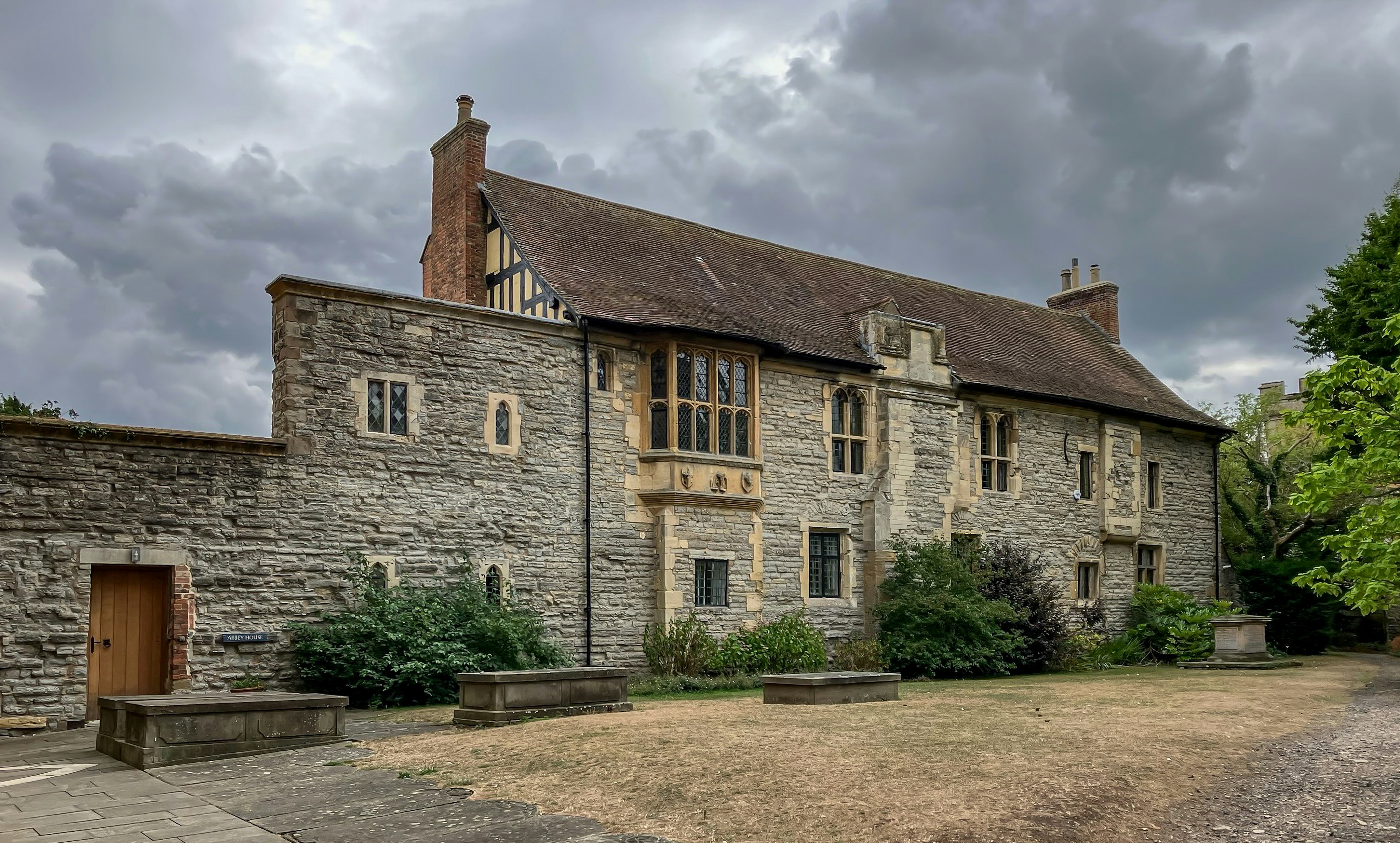Historic stone building with a wooden upper section, surrounded by greenery and a cloudy sky.
