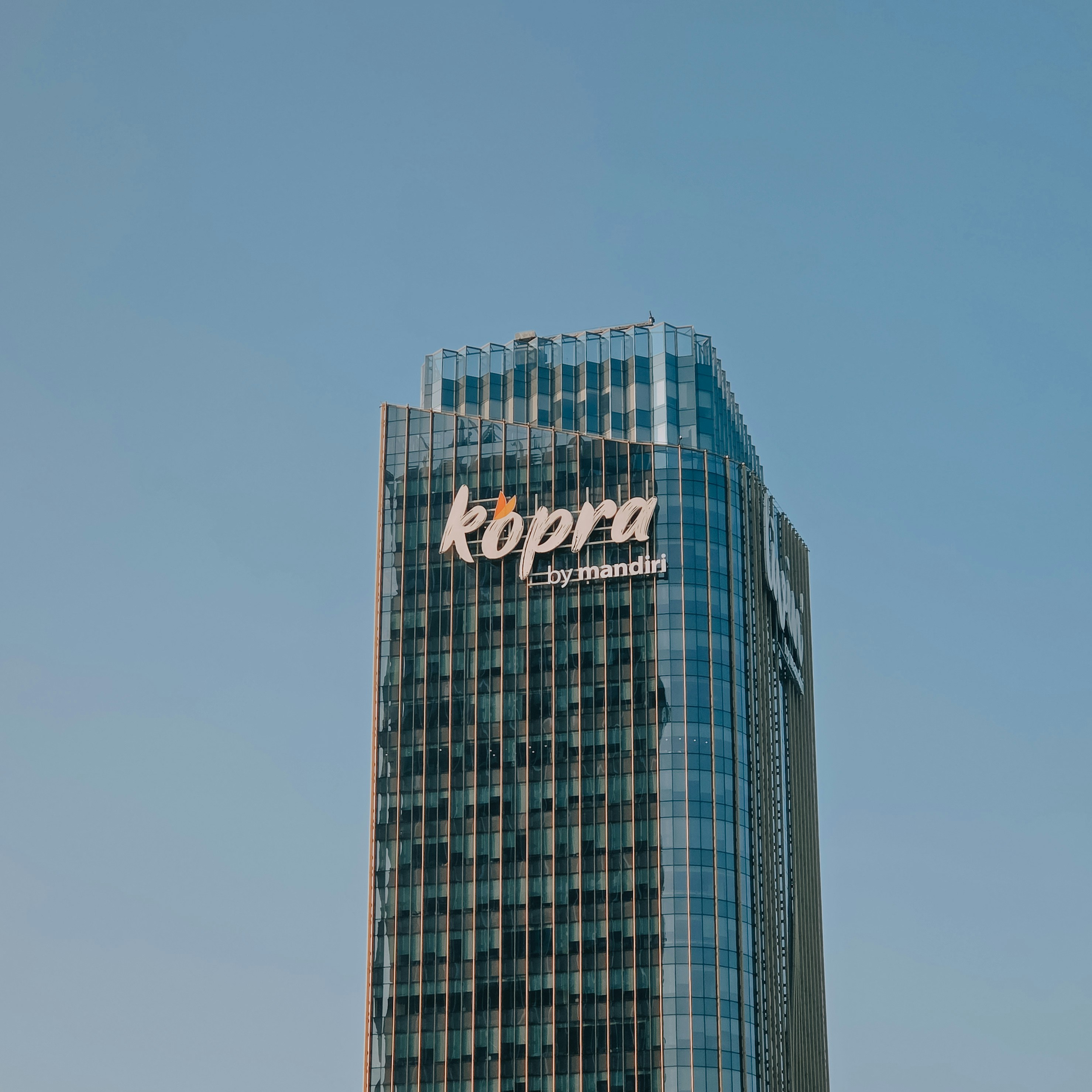 Kopra by Mandiri building | A modern skyscraper is shown under a blue sky.