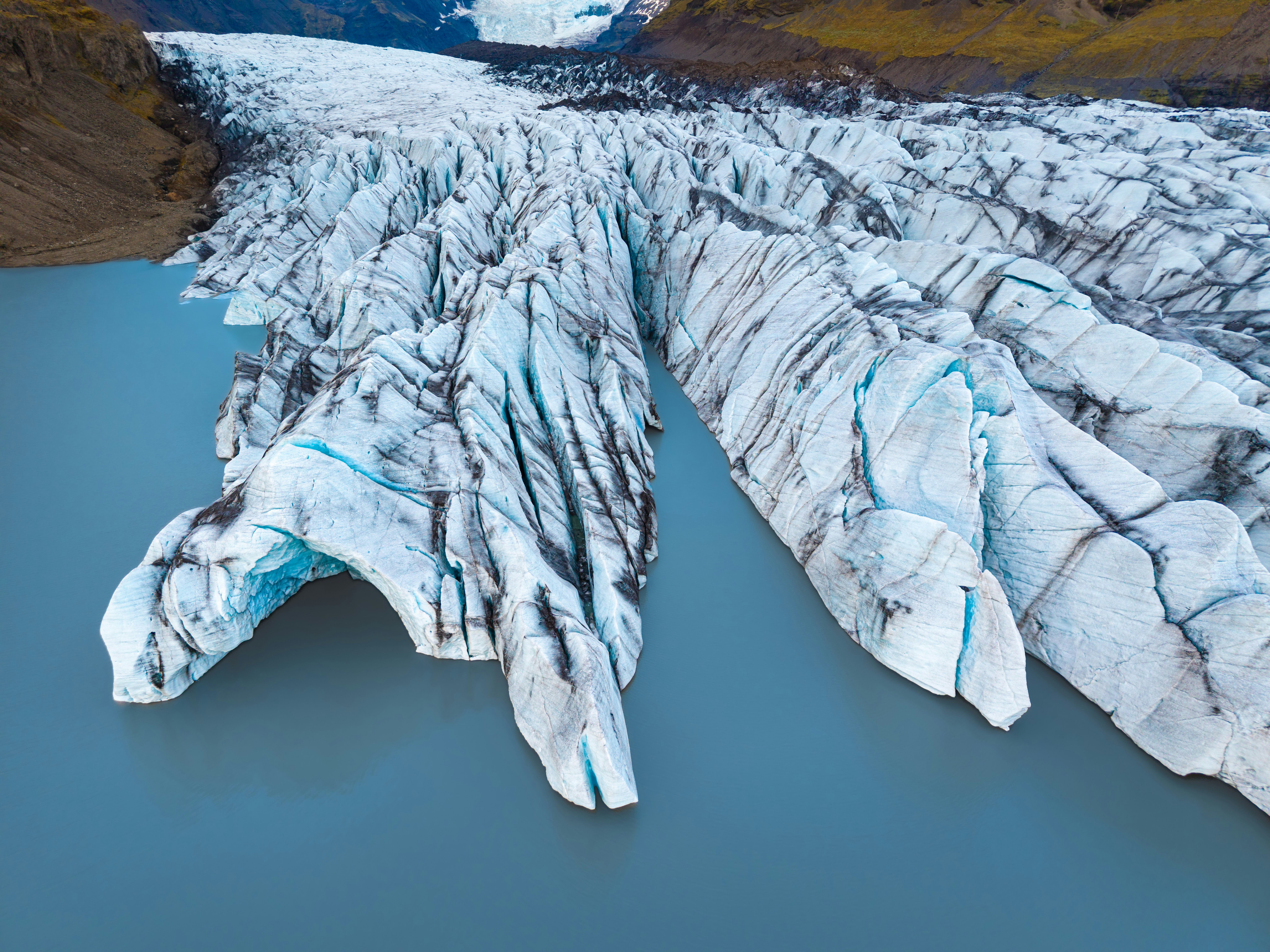 Majestic glacier flows into a serene blue lake, showcasing intricate ice formations and textures. Aerial view reveals the dynamic interplay between ice and water.