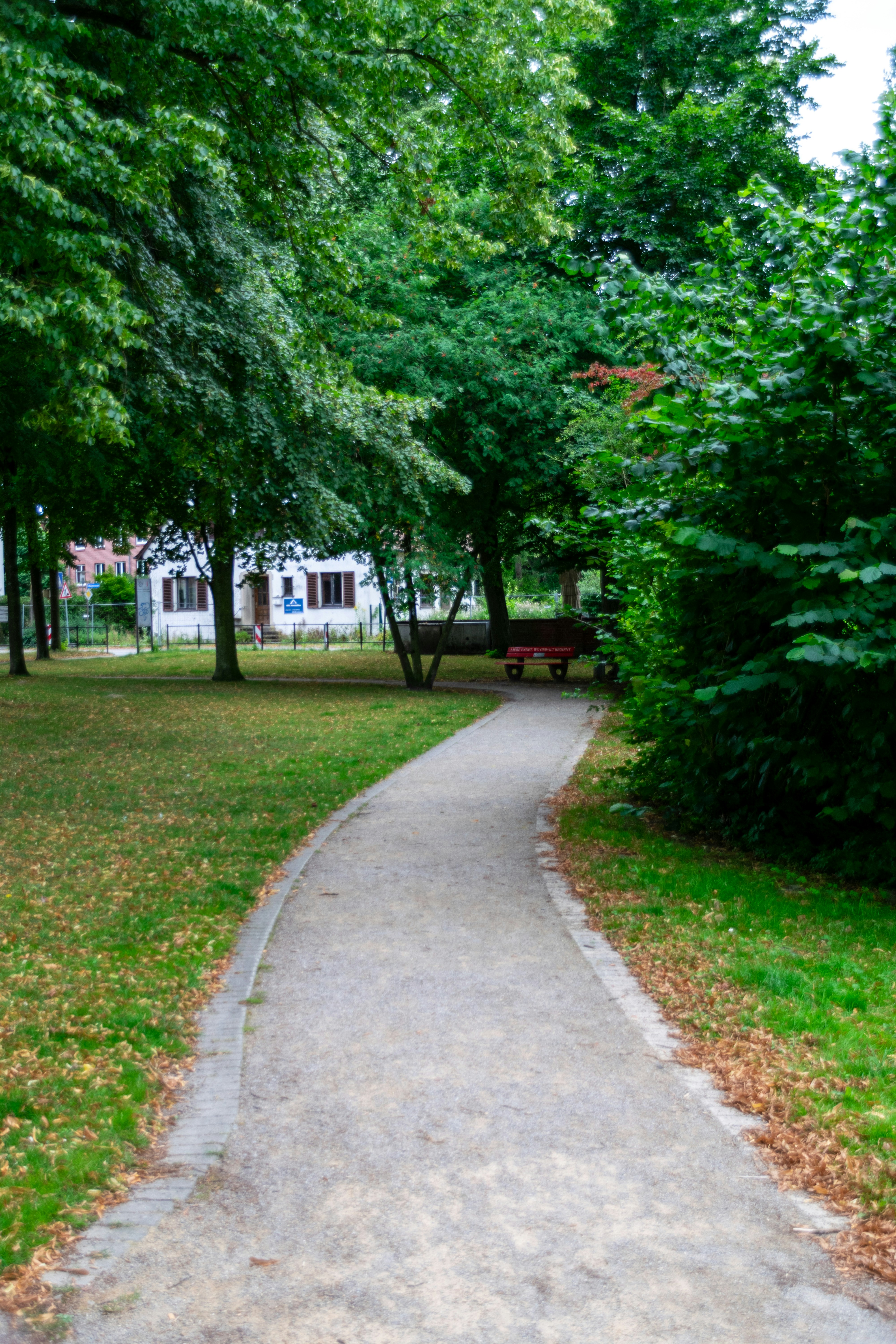 Winding gravel path meandering through a lush green park, bordered by trees and shrubs. A hint of a wooden cart is visible in the background.