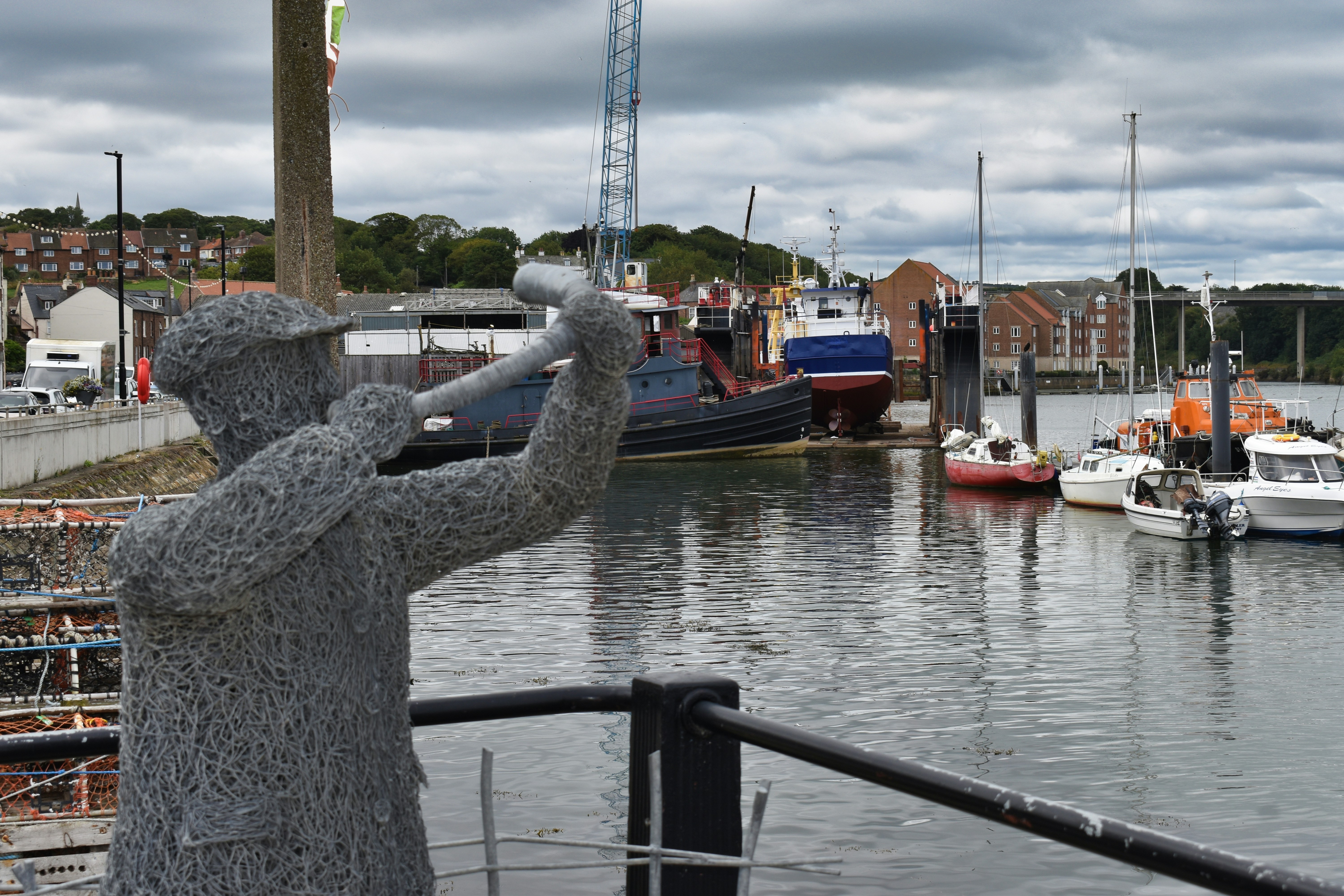 Wire sculpture of a fisherman signaling from the dock, overlooking a harbor filled with boats and a cloudy sky.