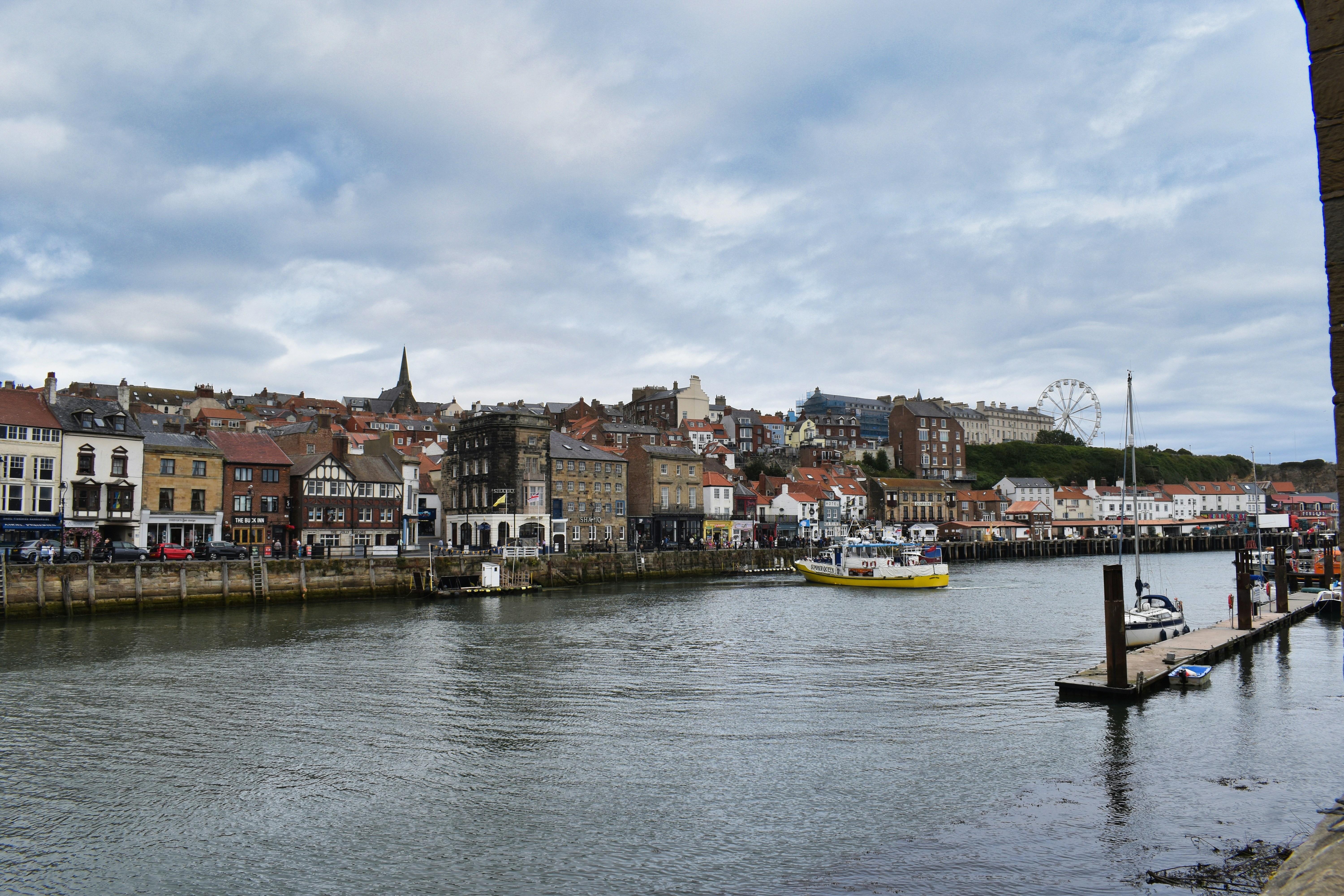 Charming seaside town with colorful buildings lining the harbor, featuring a small boat and a ferris wheel in the background.