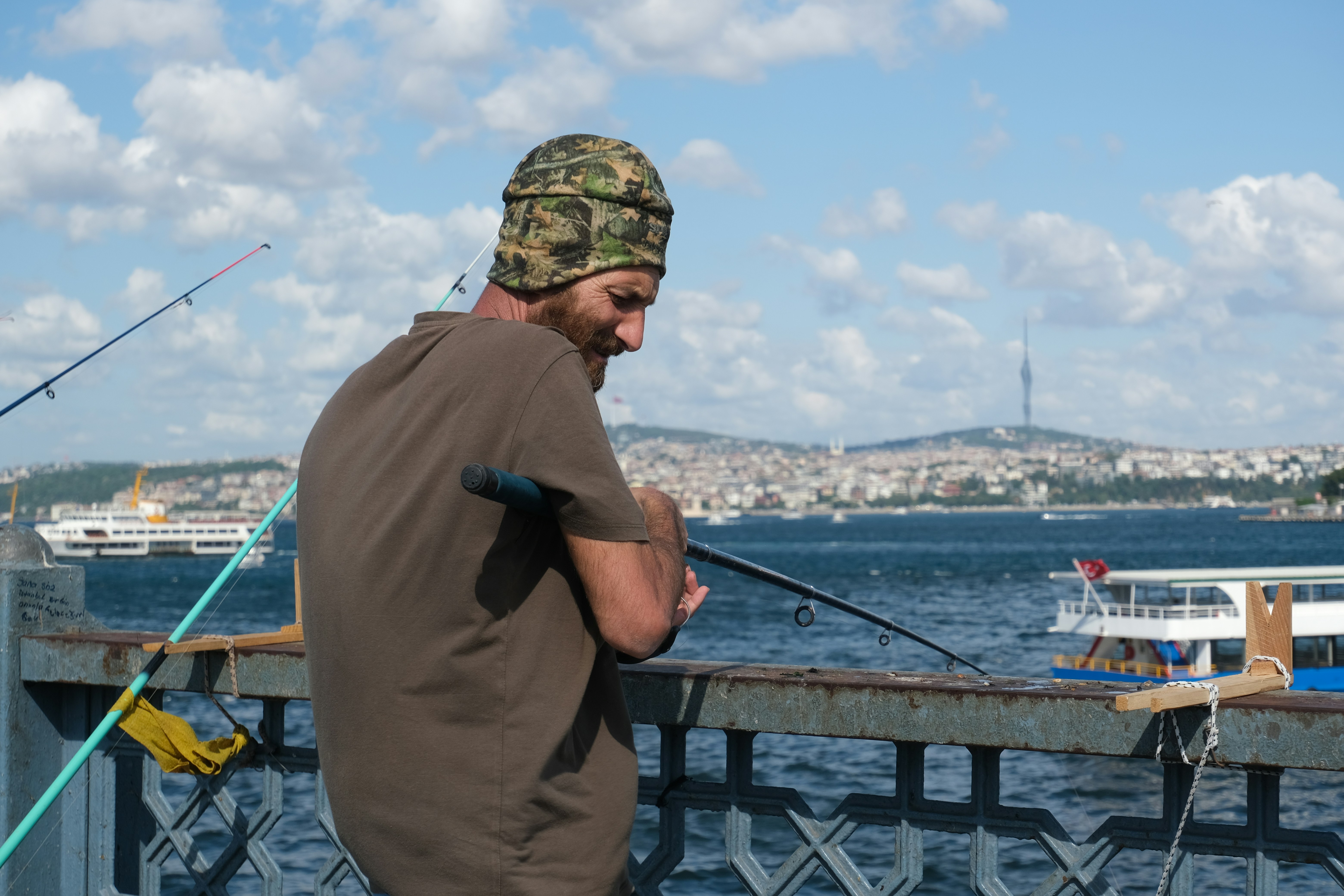 Fisherman adjusting his fishing rod on a bustling pier in Istanbul, with the city skyline and boats in the background.