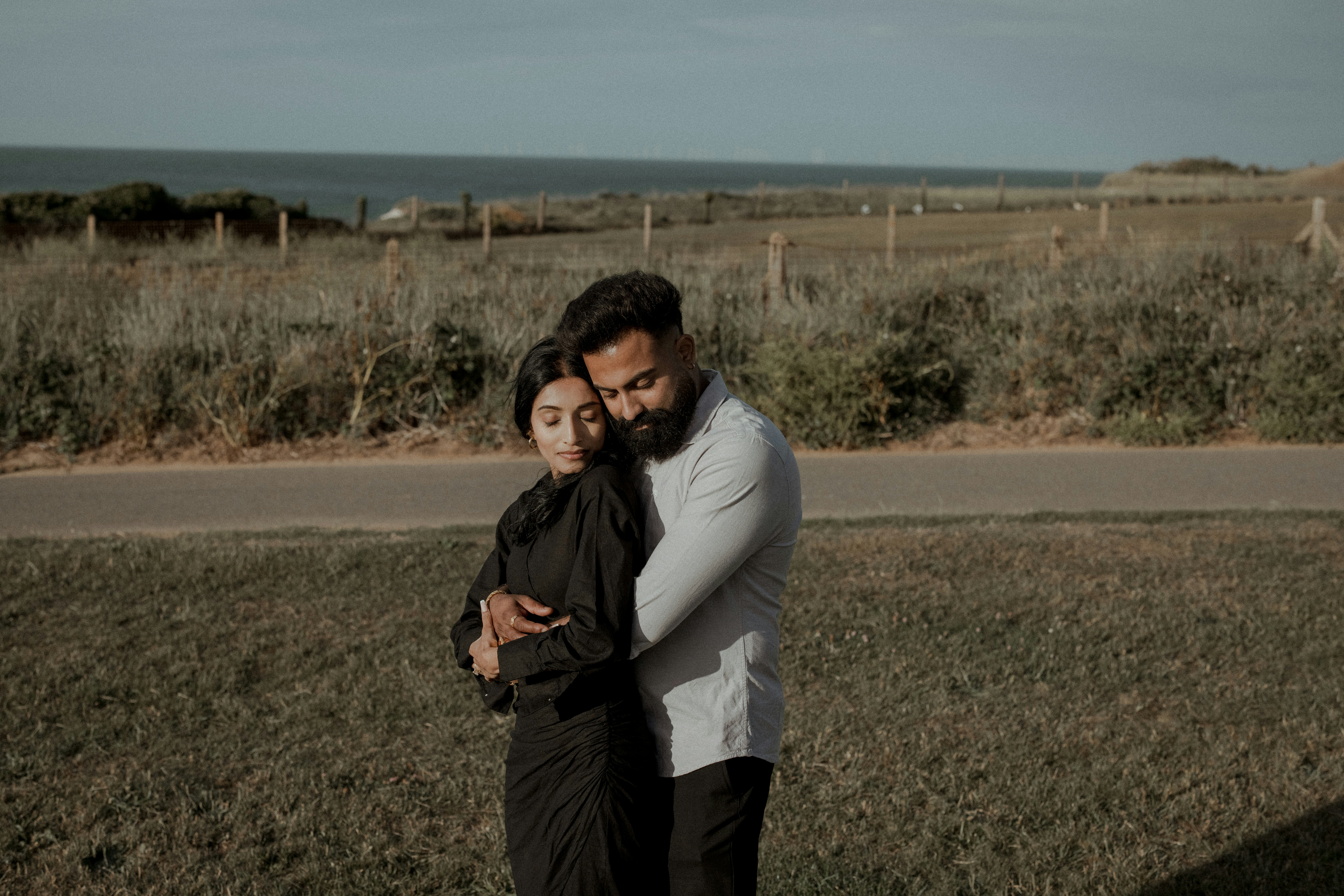 Couple embracing in a serene outdoor setting, with a lush green field and ocean backdrop. The warmth of their connection contrasts with the cool tones of the landscape.
