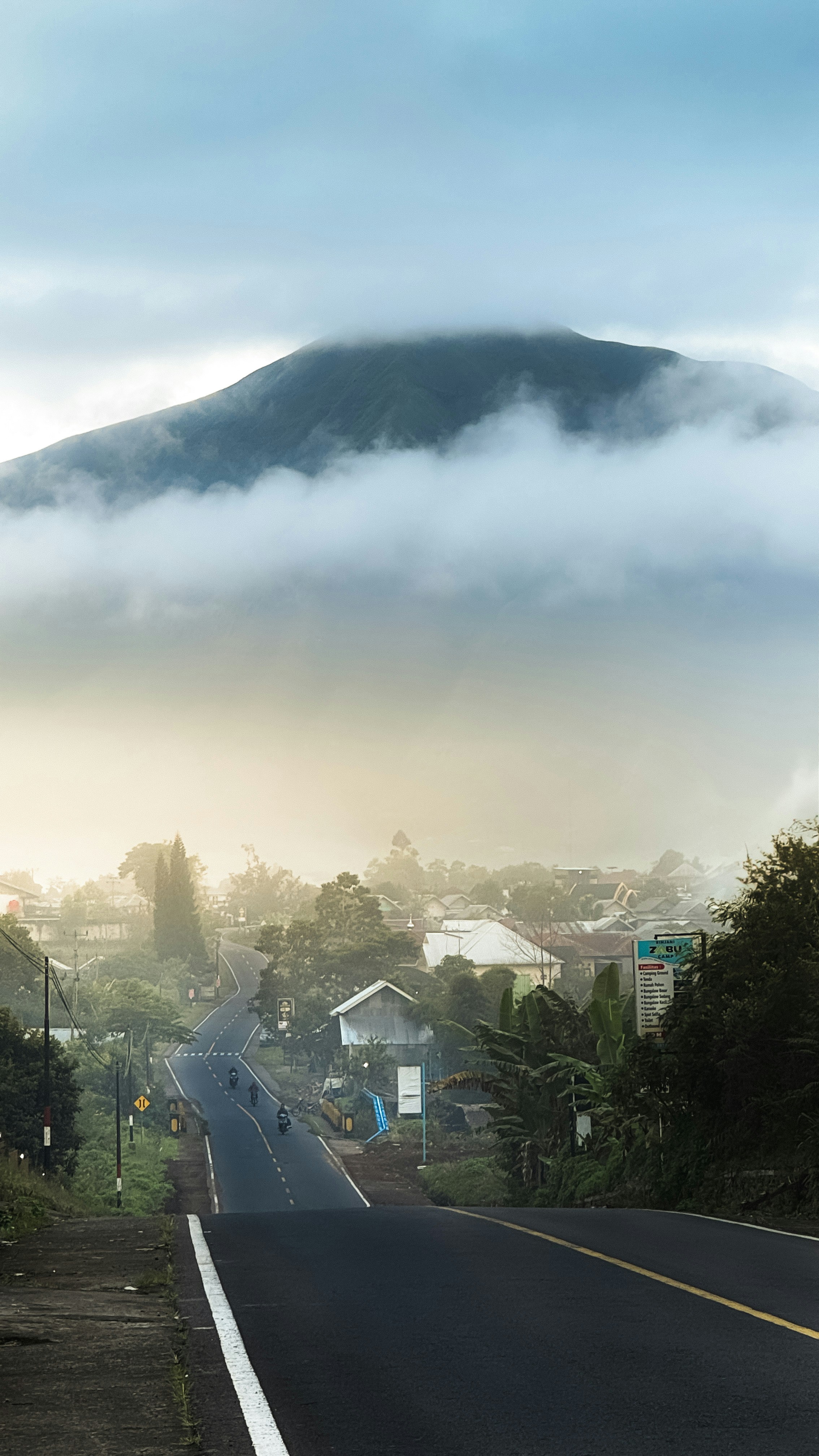 A winding road leads through a misty landscape, with a mountain partially shrouded in clouds in the background. The scene evokes a serene and tranquil atmosphere.