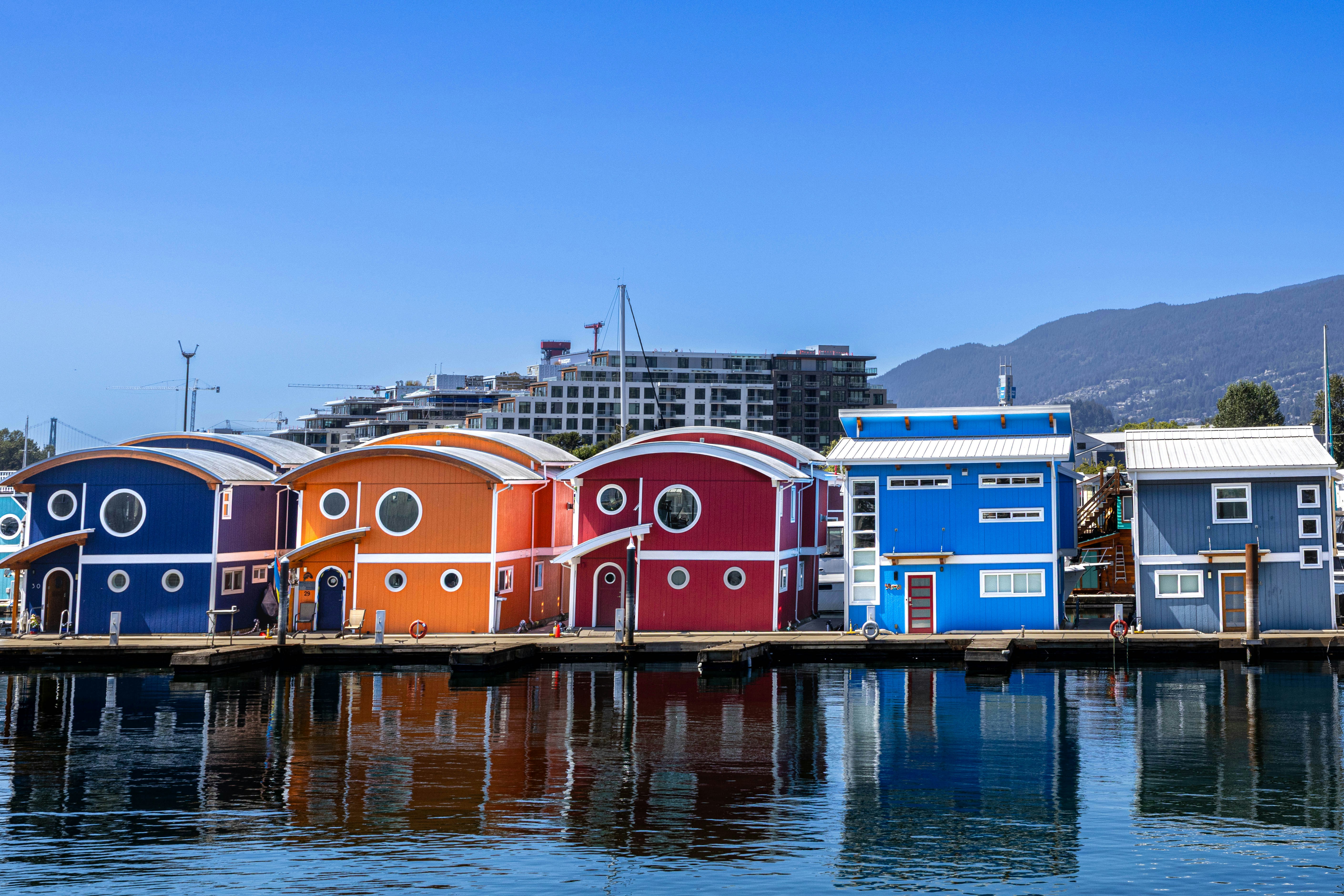 Row of vividly painted floating homes moored at a North Vancouver marina: circular porthole windows, bold primary colours and Canadian flags mirror crisply in the still harbour water beneath a clear blue Pacific Northwest sky, with distant mountains and city buildings framing the coastal scene. | Colorful floating houses line a sunny waterfront.
