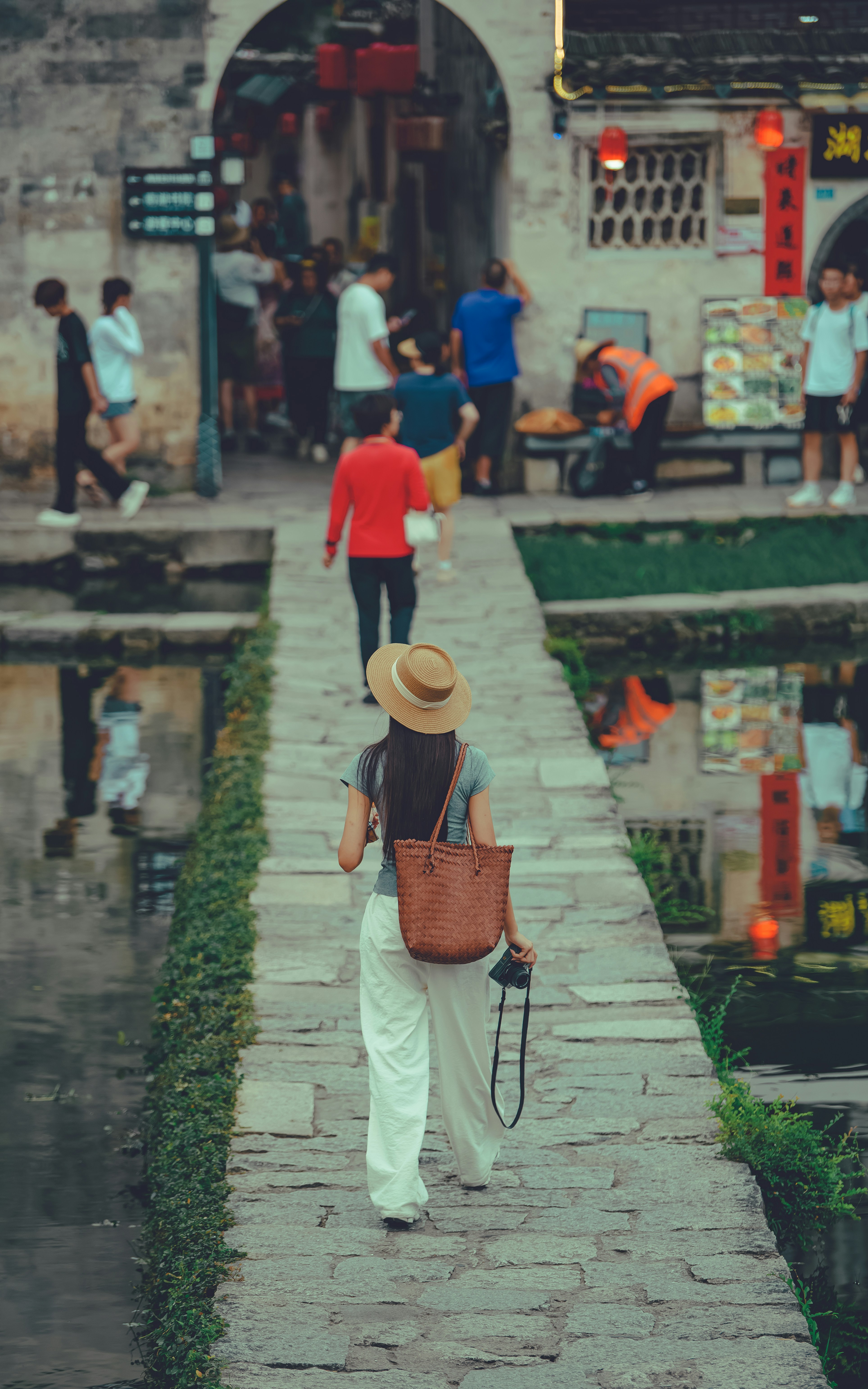 A woman in a straw hat walks along a stone path beside a reflective waterway, surrounded by bustling market activity in a quaint village.