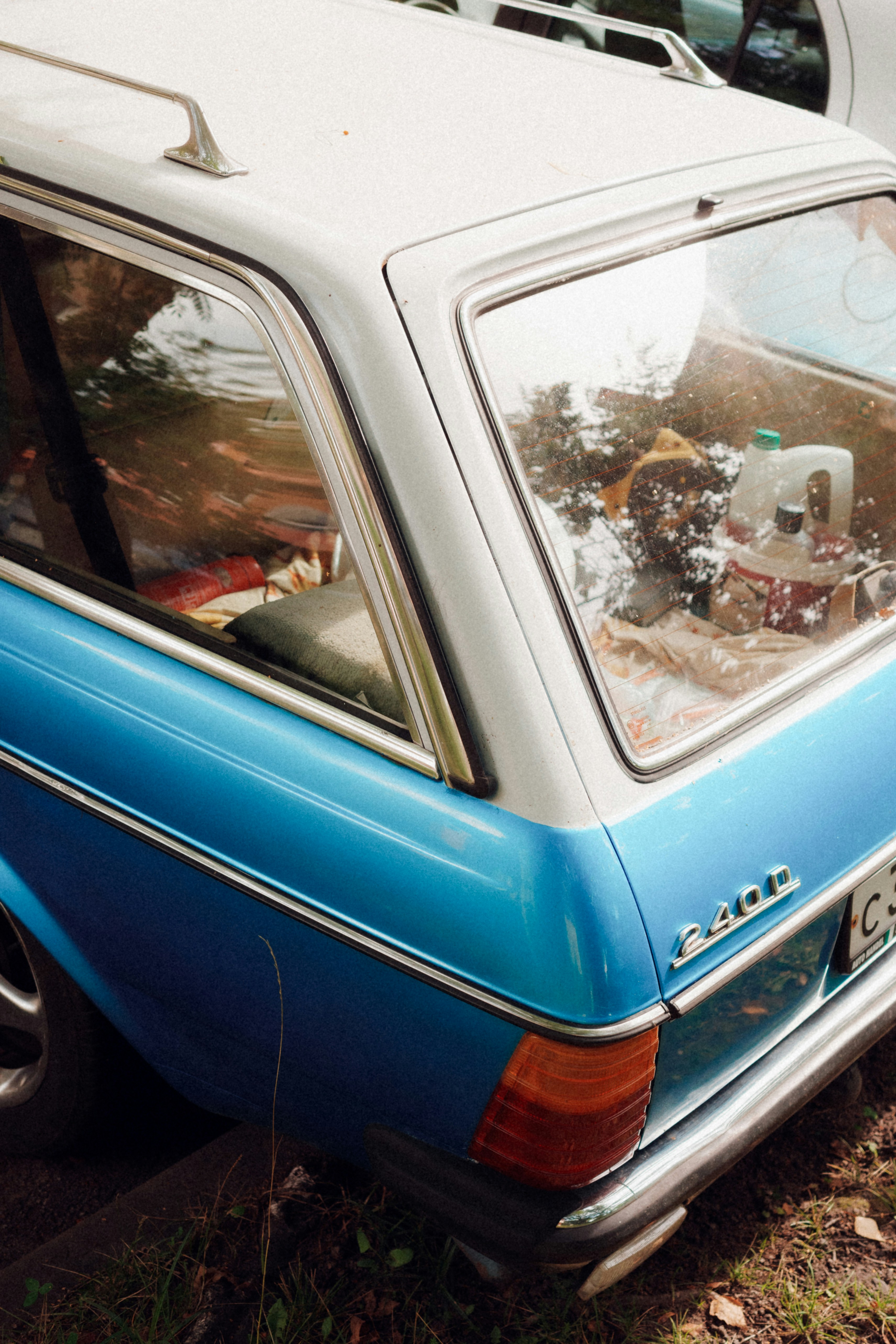 An old blue car parked amidst nature, showcasing its rear view with reflections in the glass and hints of foliage inside.