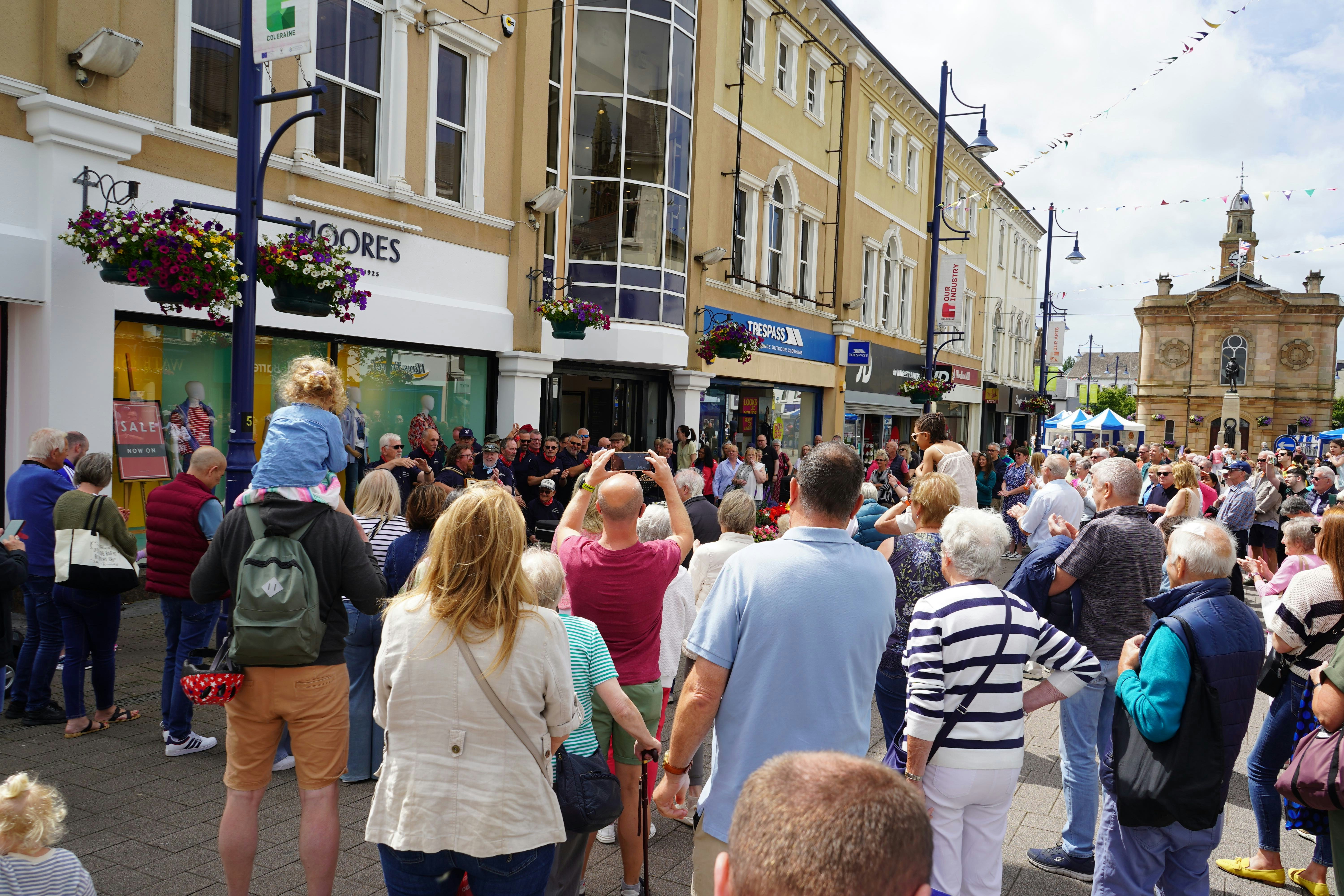 Crowd of people gathered in a lively town square, engaging with a performance or event. Brightly colored storefronts and festive decorations create a vibrant atmosphere.