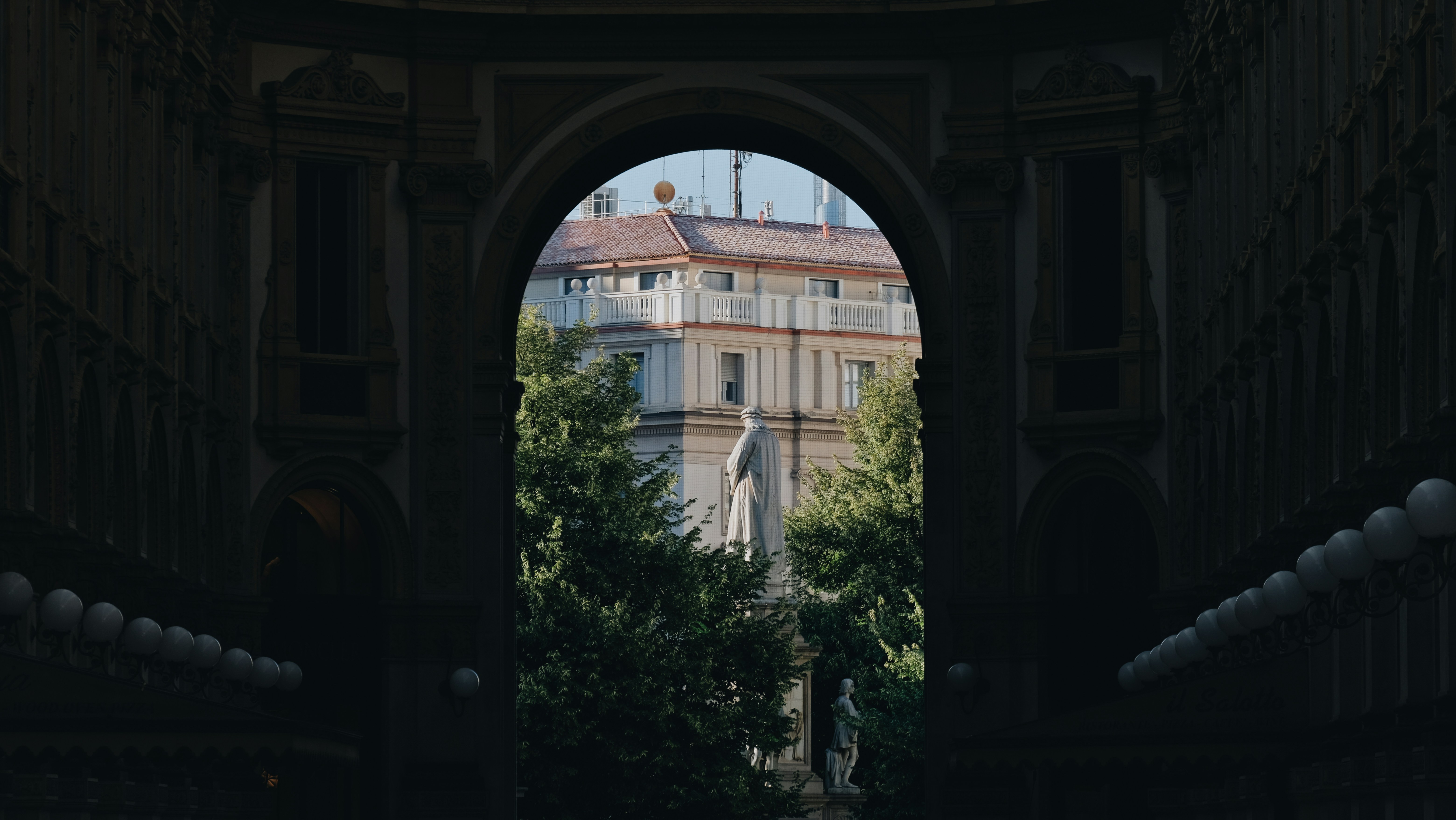 A view through an architectural arch, showcasing a statue and greenery beyond, framed by contrasting shadows. The scene captures a moment of serenity in a bustling environment.