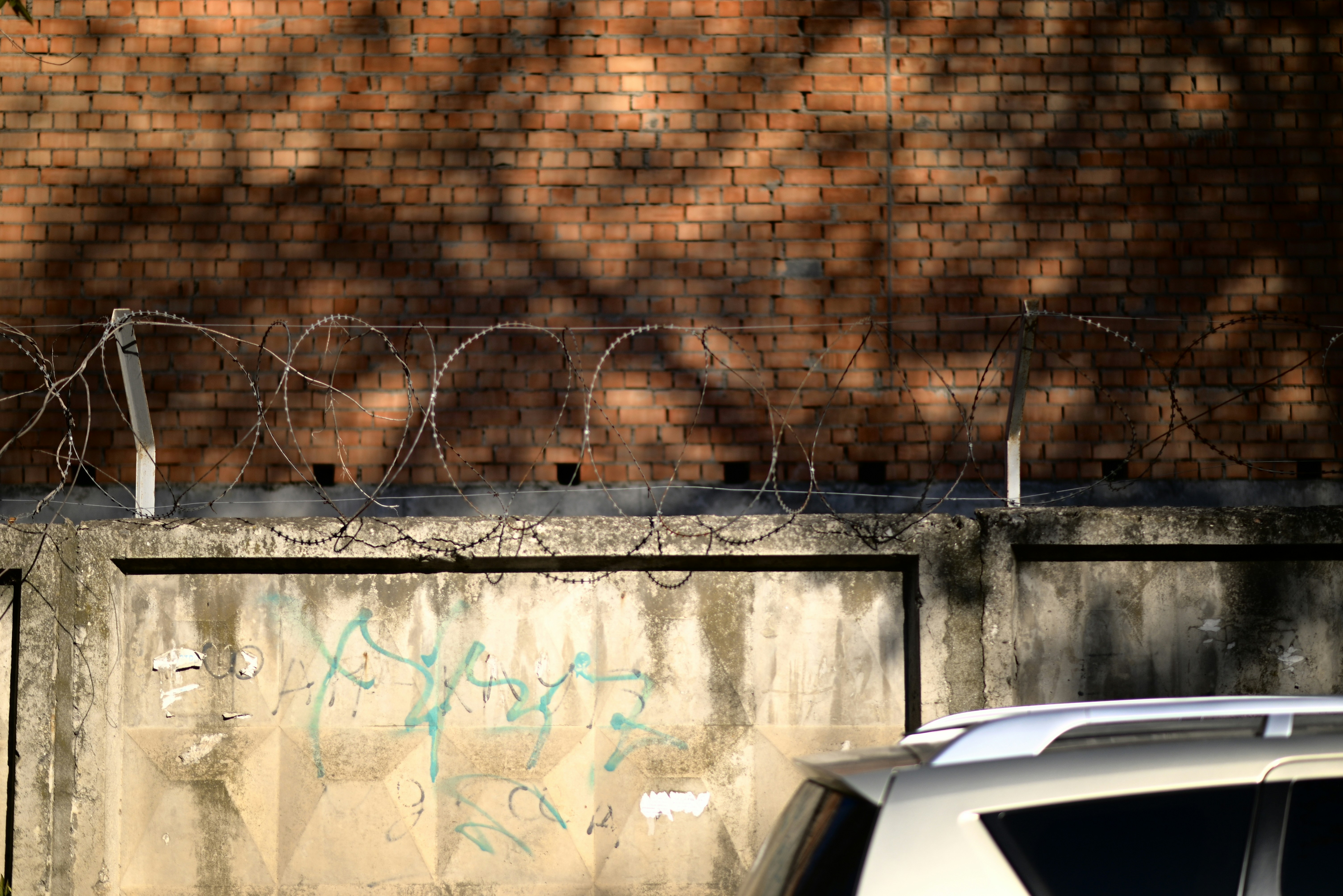 Barbed wire atop a weathered concrete wall, with graffiti partially visible, set against a backdrop of textured brickwork. The scene captures the intersection of urban decay and security.