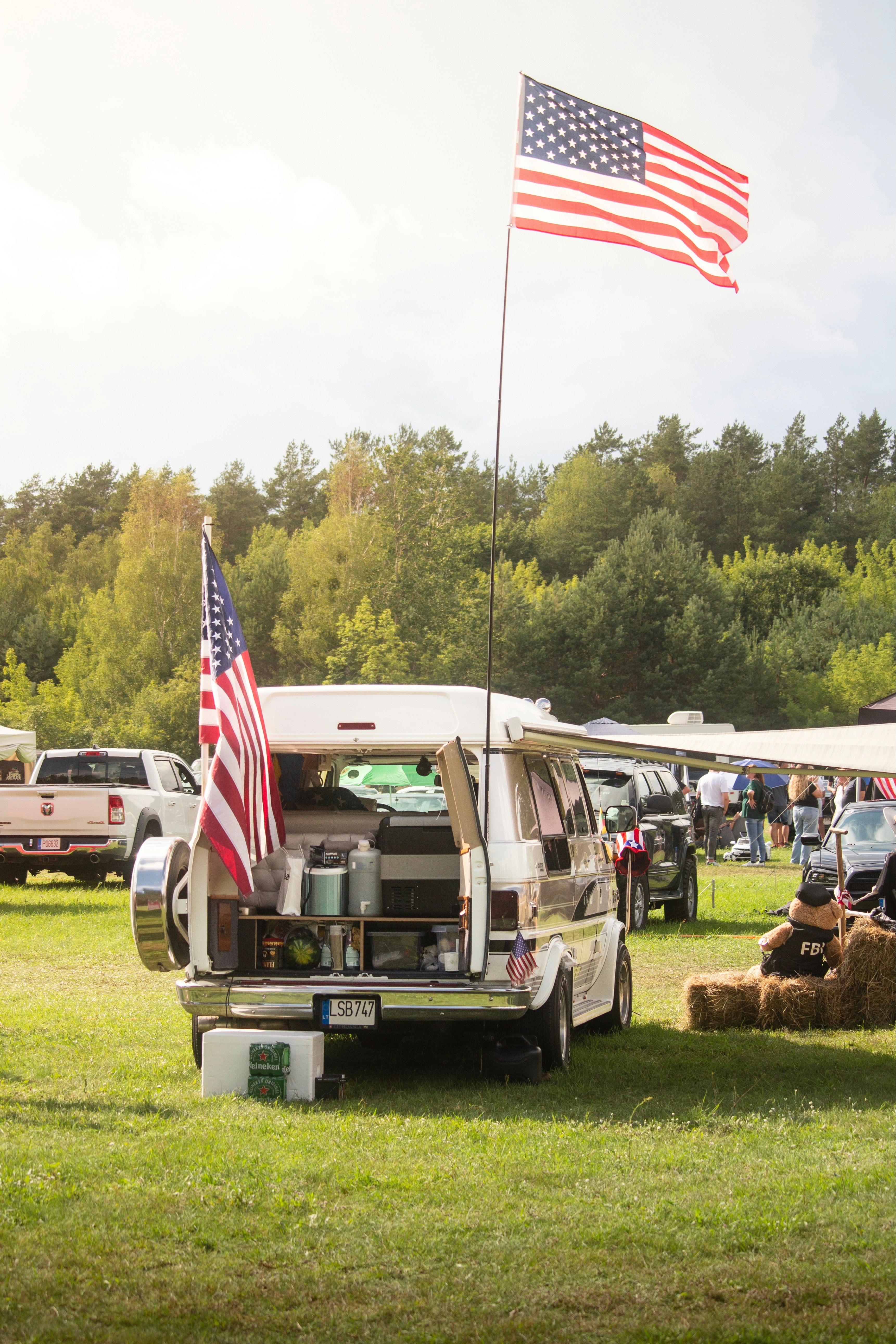 A vintage van adorned with American flags, parked in a grassy field, surrounded by fellow festival-goers enjoying the outdoors.