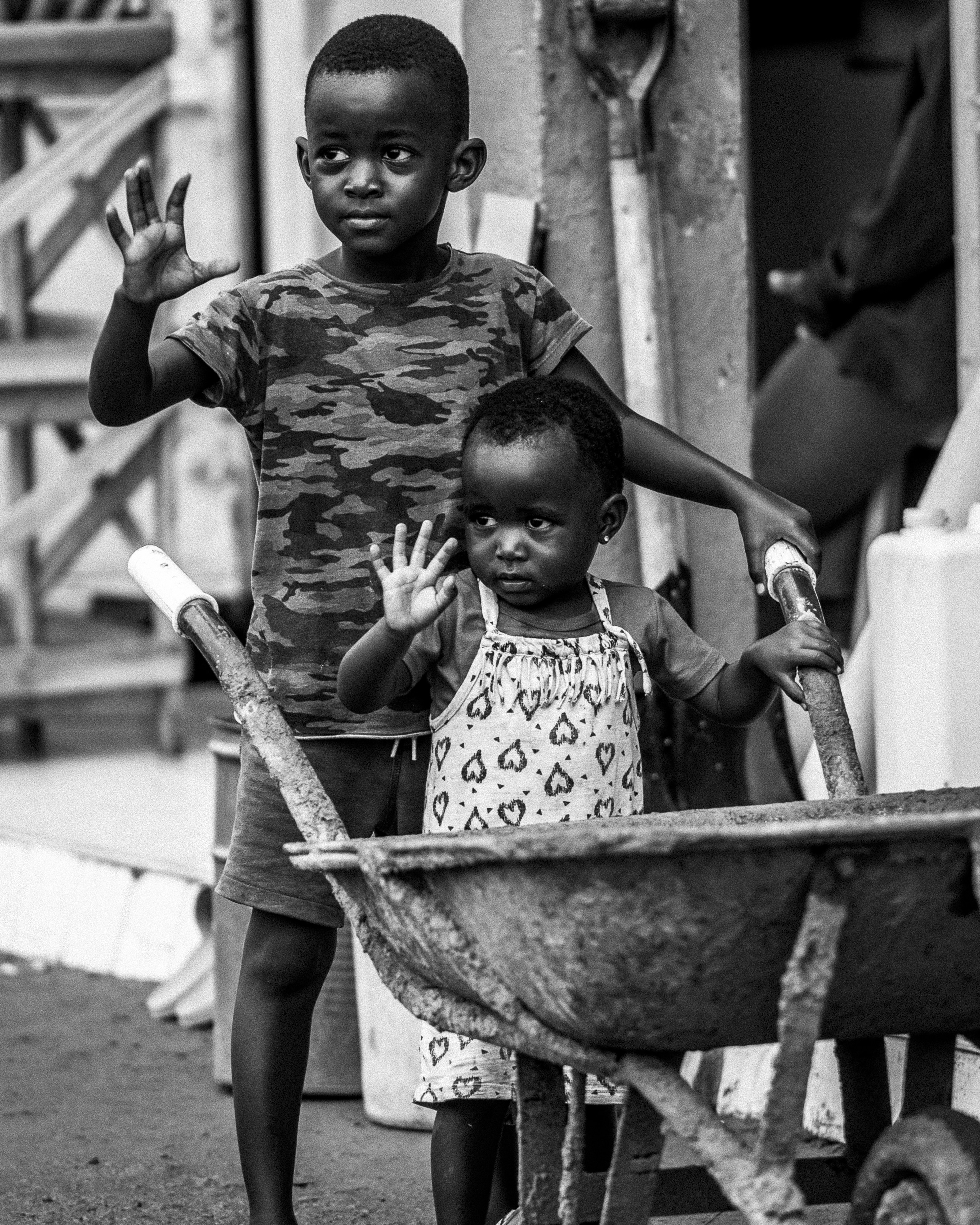 Two children waving cheerfully beside a wheelbarrow, embodying innocence and playfulness in a lively environment.