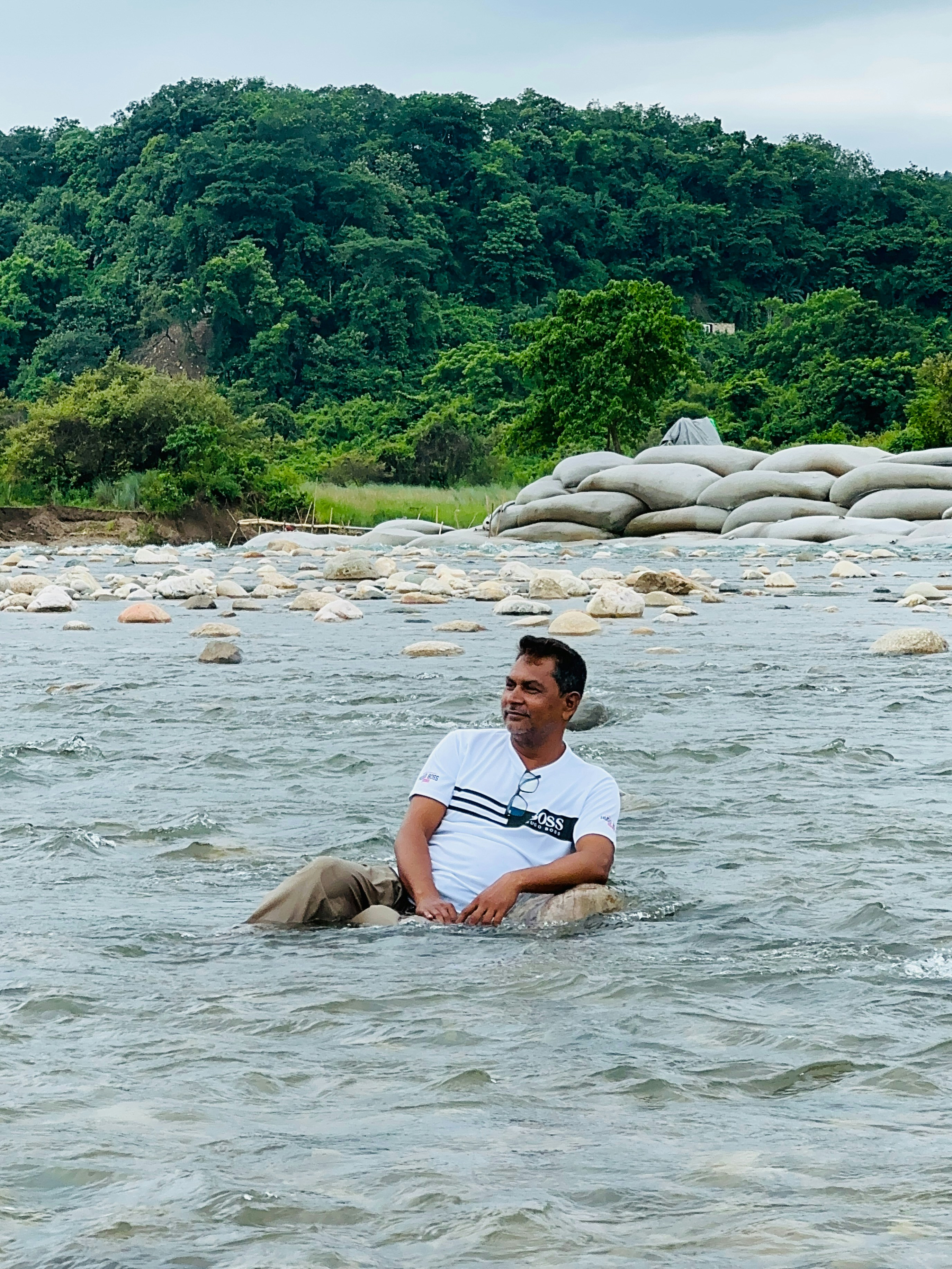 Man resting on a partially submerged rock amid a shallow river with lush green hills in the background