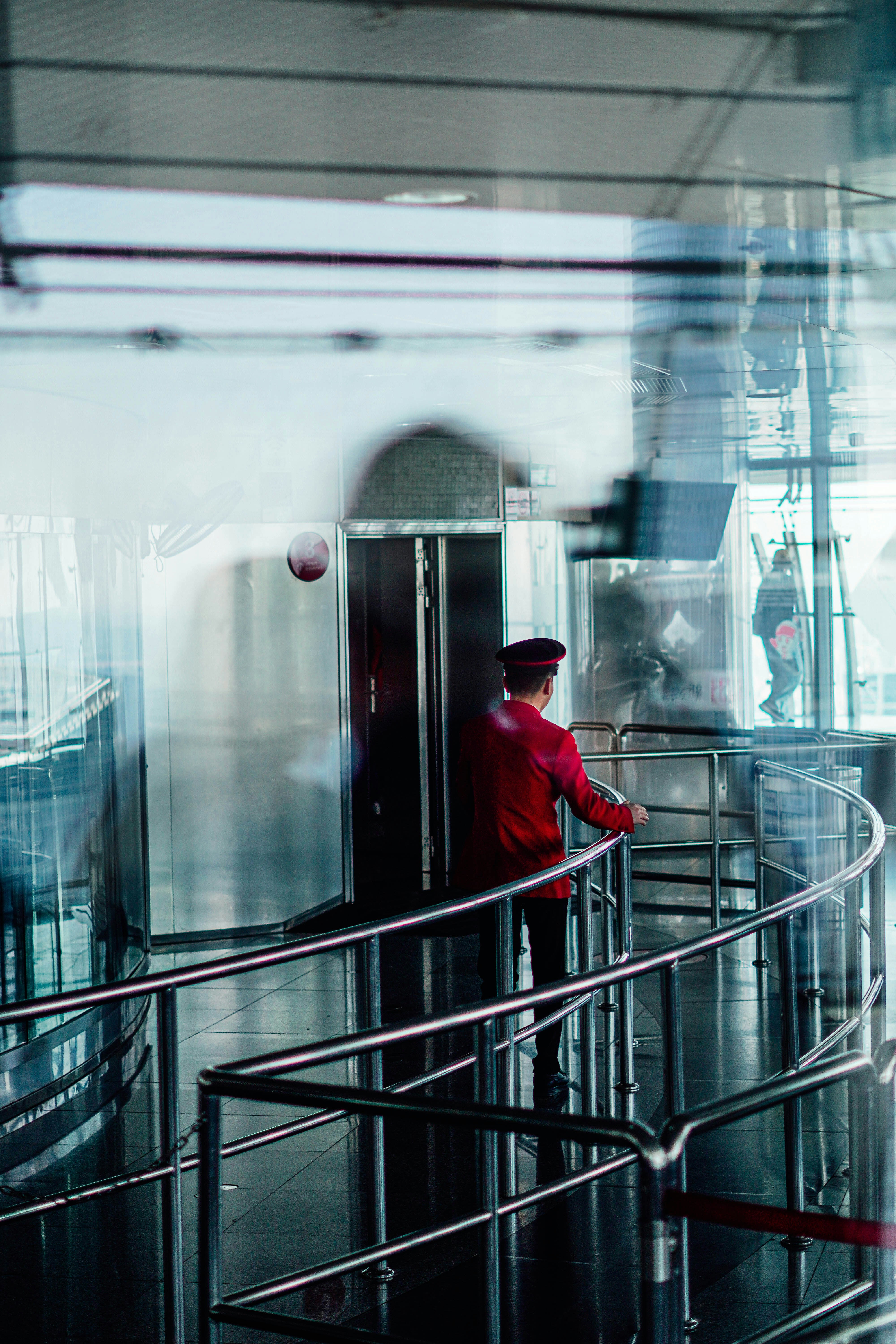 A figure in a red uniform stands at a curved railing in a modern observation area, surrounded by reflections and glass. The scene captures a moment of stillness amidst the architectural elegance.