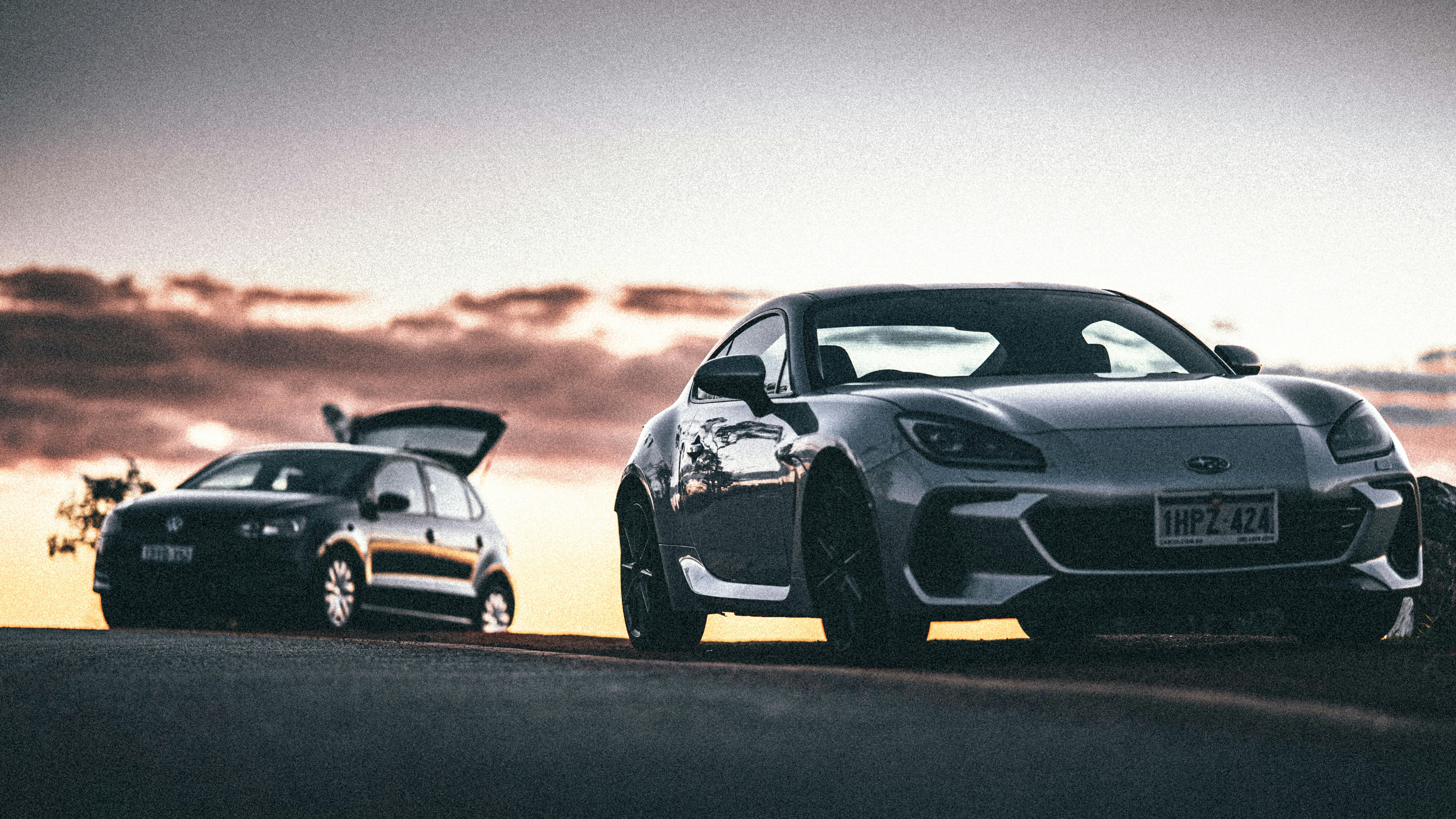 Two cars parked on a scenic overlook during twilight, with one car prominently featured in the foreground and the other in the background under a colorful sky.