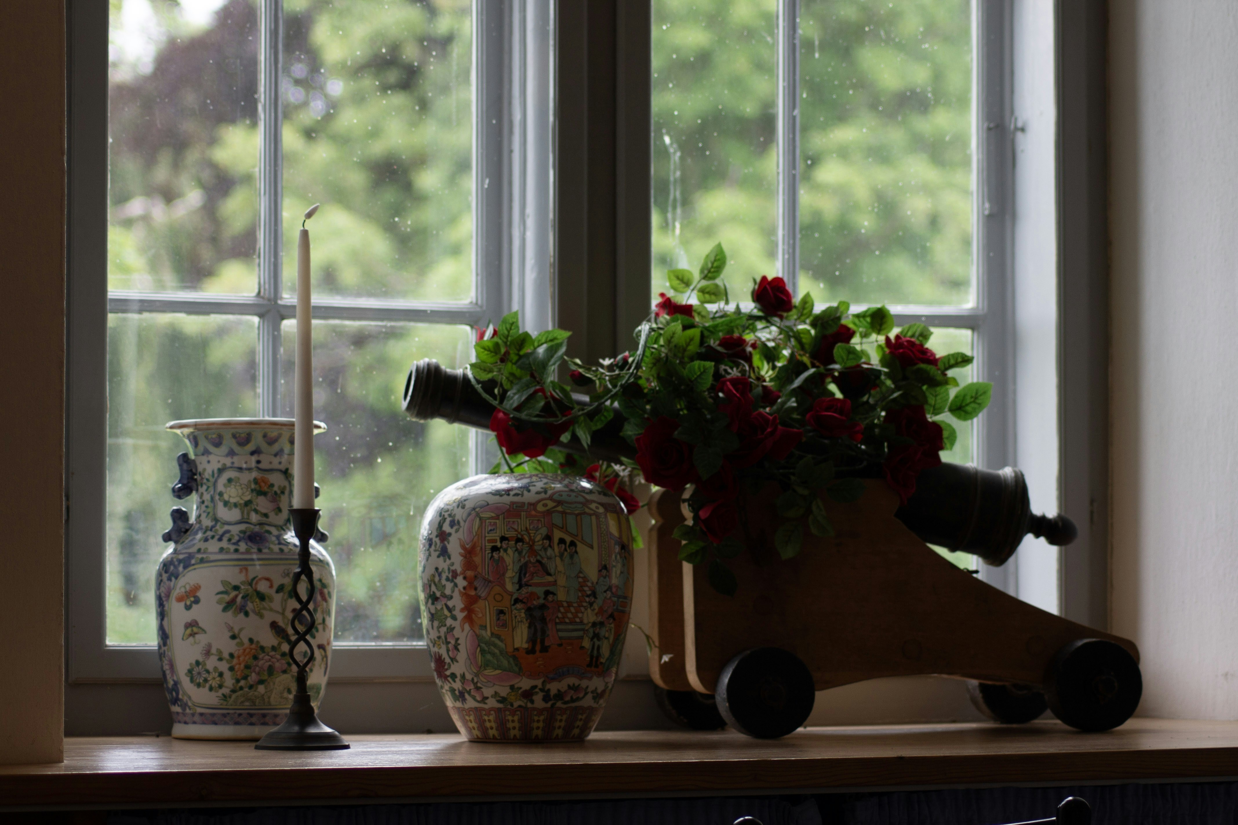 A charming arrangement of a decorative vase, a wooden cannon planter filled with red roses, and a candle holder, all set against a window backdrop.