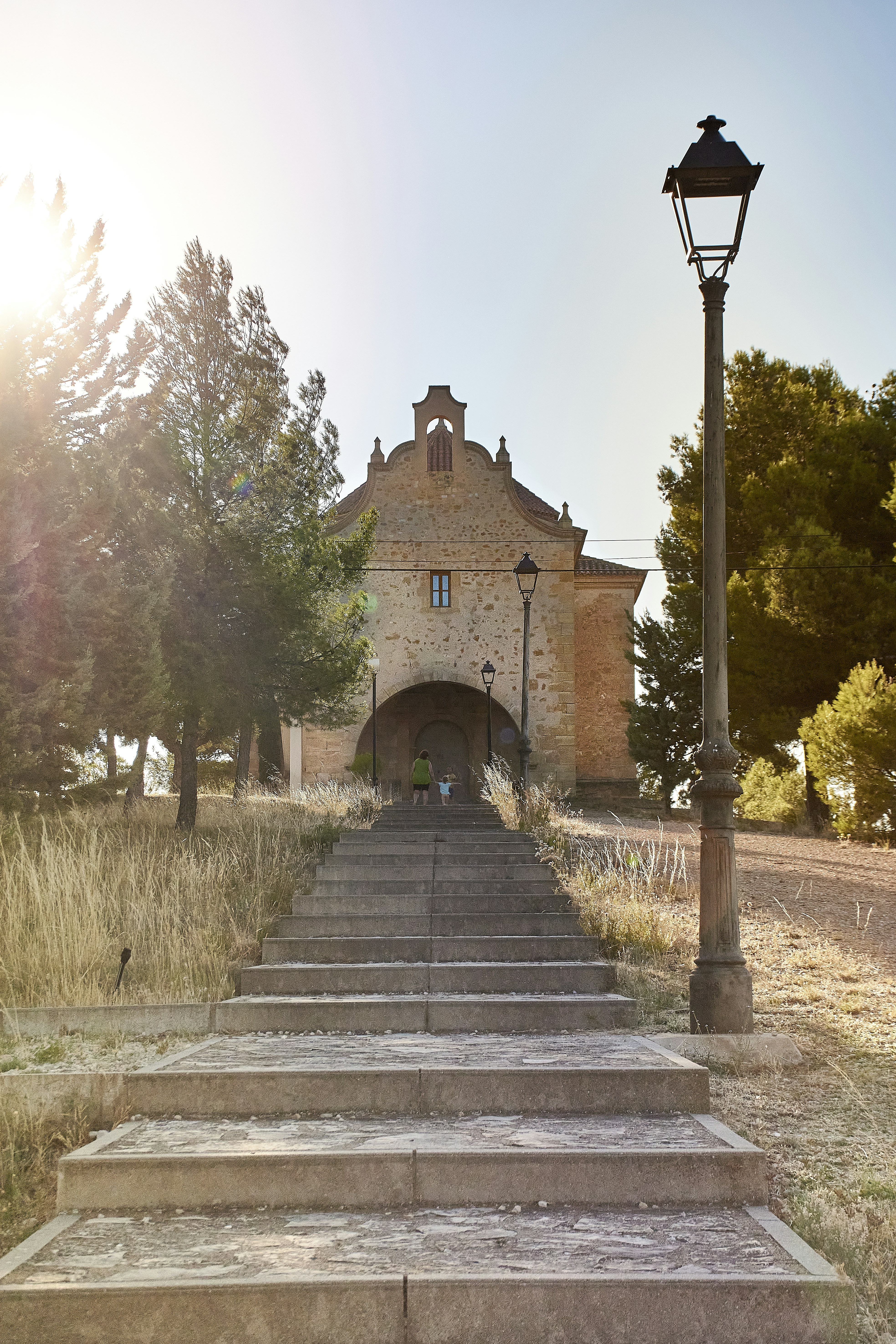 Stone steps leading up to a historic chapel surrounded by trees and lampposts, illuminated by soft sunlight.