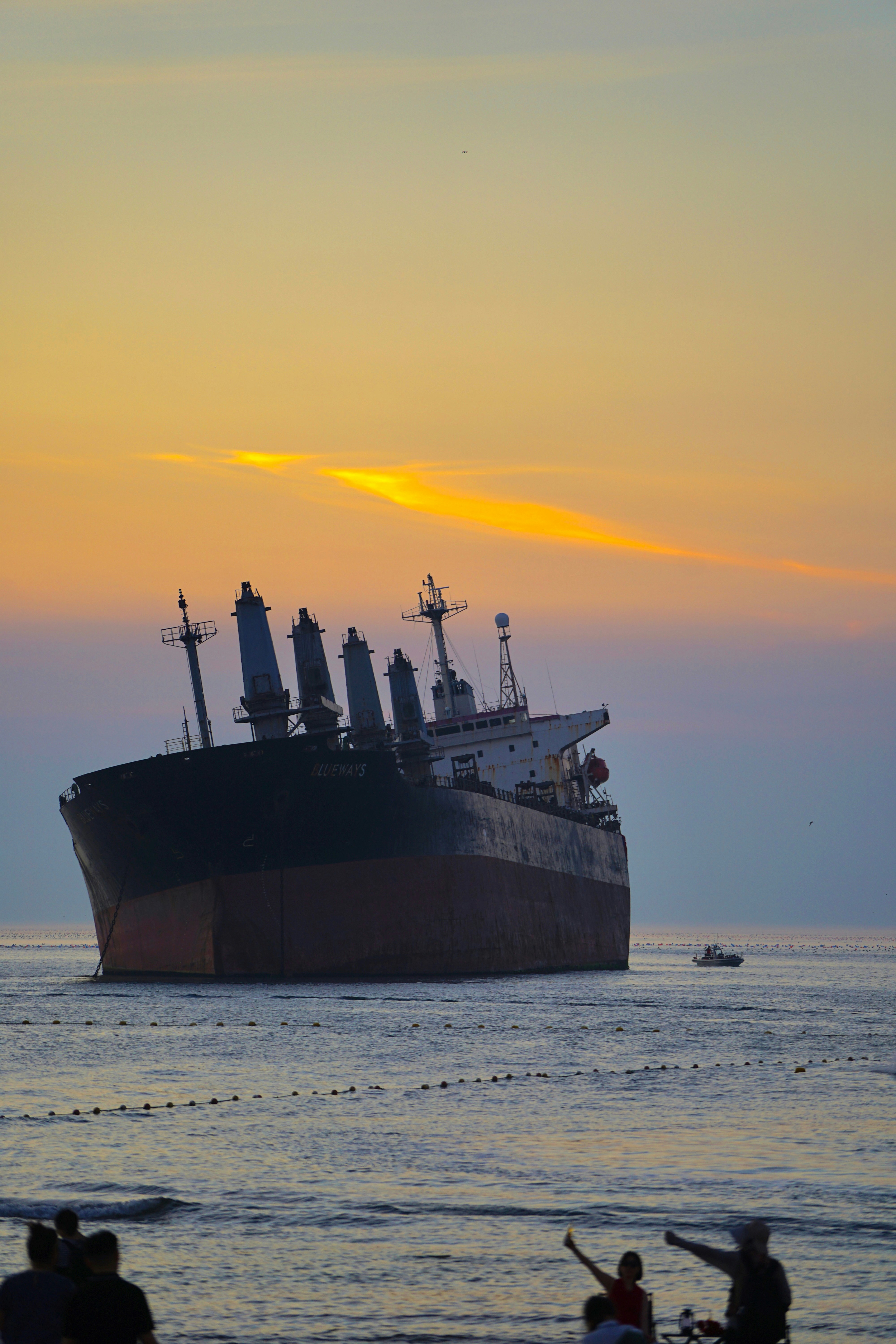 Large cargo ship anchored off the coast during a vibrant sunset, with silhouettes of people enjoying the beach in the foreground.