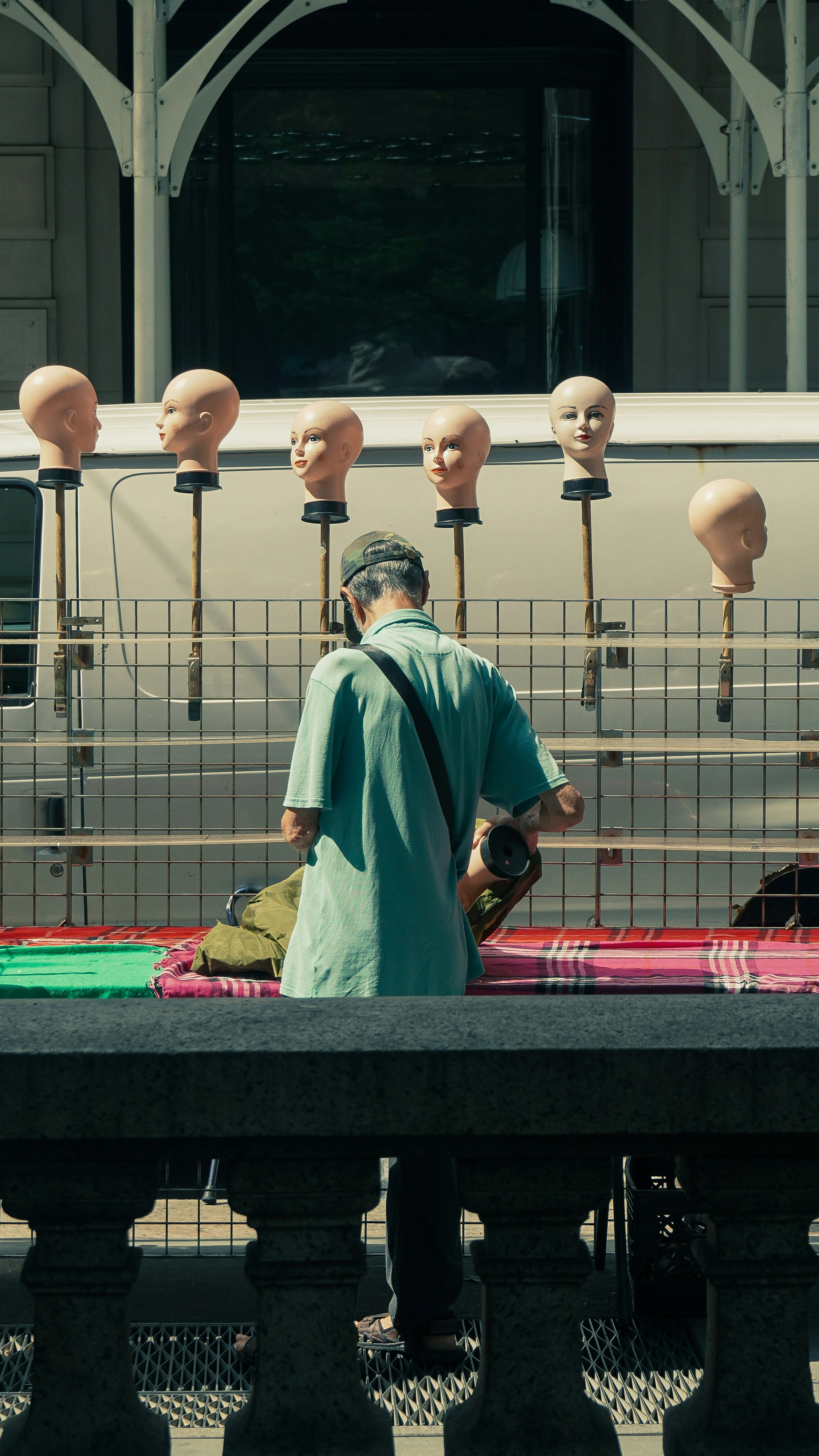 A man in a teal shirt examines items on a colorful table, with a row of mannequin heads observing from behind a fence. 