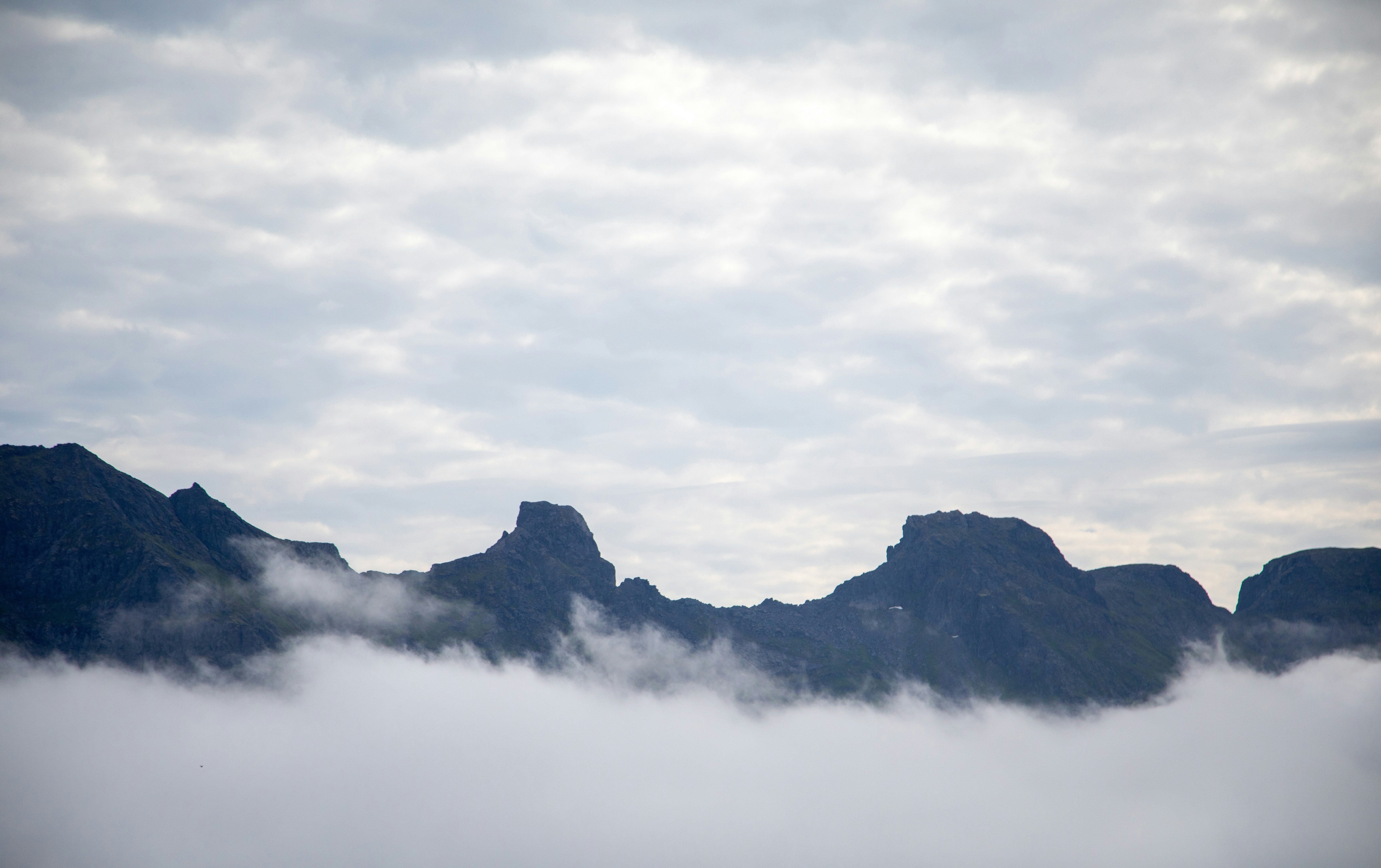 Dramatic mountain silhouette emerging from a blanket of clouds under an overcast sky.