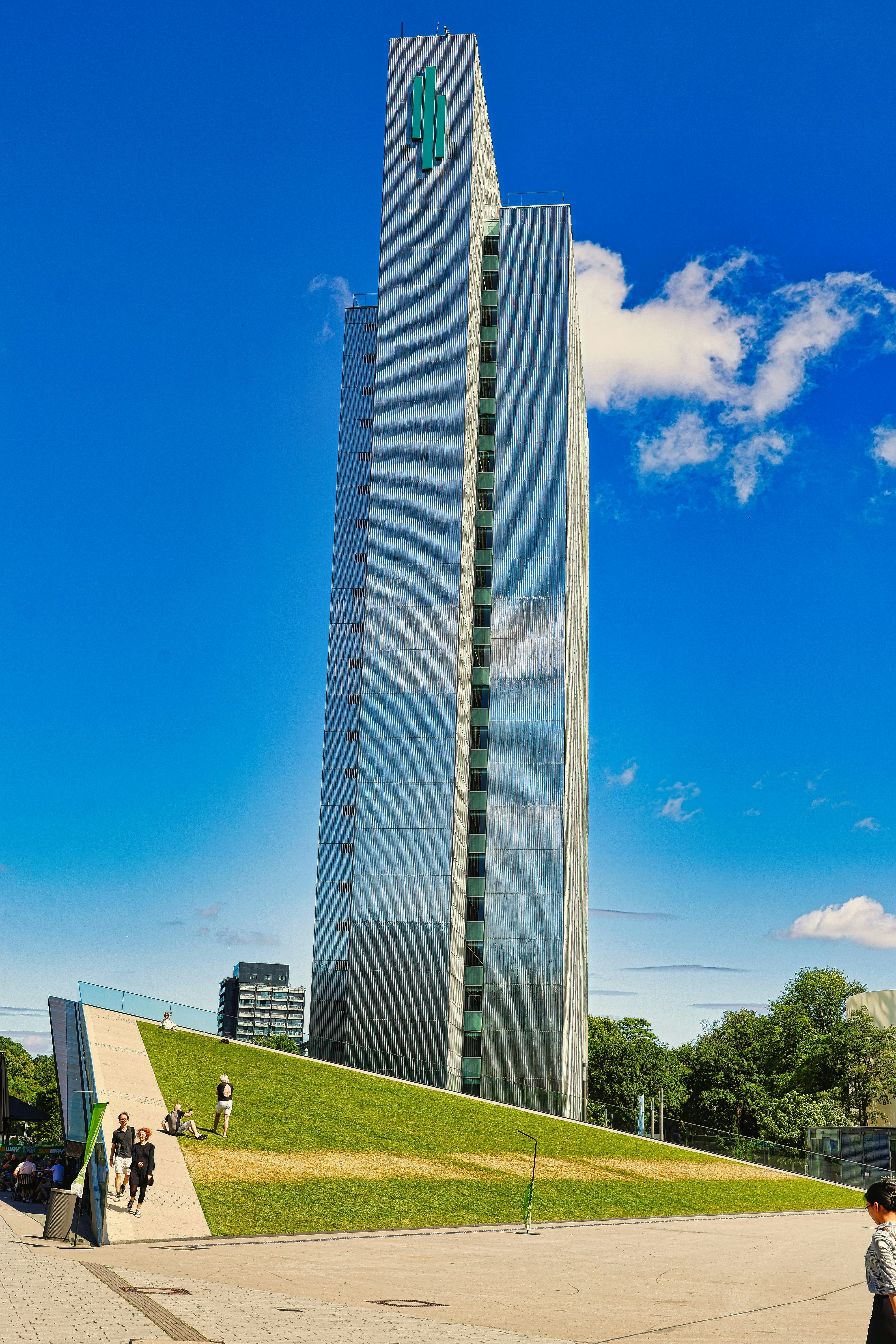 La Schadowstraße, au cœur de Düsseldorf, illustre une approche exemplaire de l’espace public dédié aux mobilités douces. Sa zone piétonne commerçante intègre une large piste cyclable centrale, bordée de généreux trottoirs ombragés pour les piétons et des espaces de stationnement pour les vélos. Accessible facilement en tram et en métro, elle constitue un modèle d’accessibilité écomobile dans un cadre animé et attractif. Rarement une rue commerçante combine avec autant d’équilibre confort piéton, circulation cycliste et transports publics. | A tall, modern building reaches towards the sky.