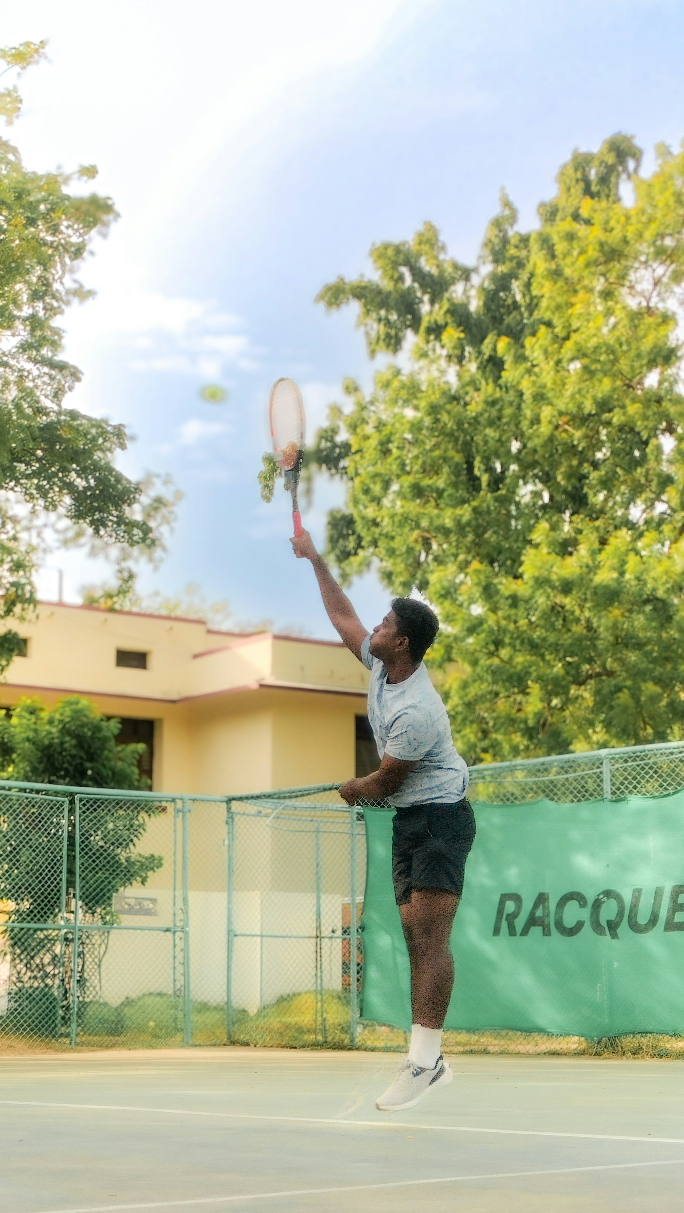 Tennis player poised to serve, with the ball in mid-air against a backdrop of greenery and a clear sky.