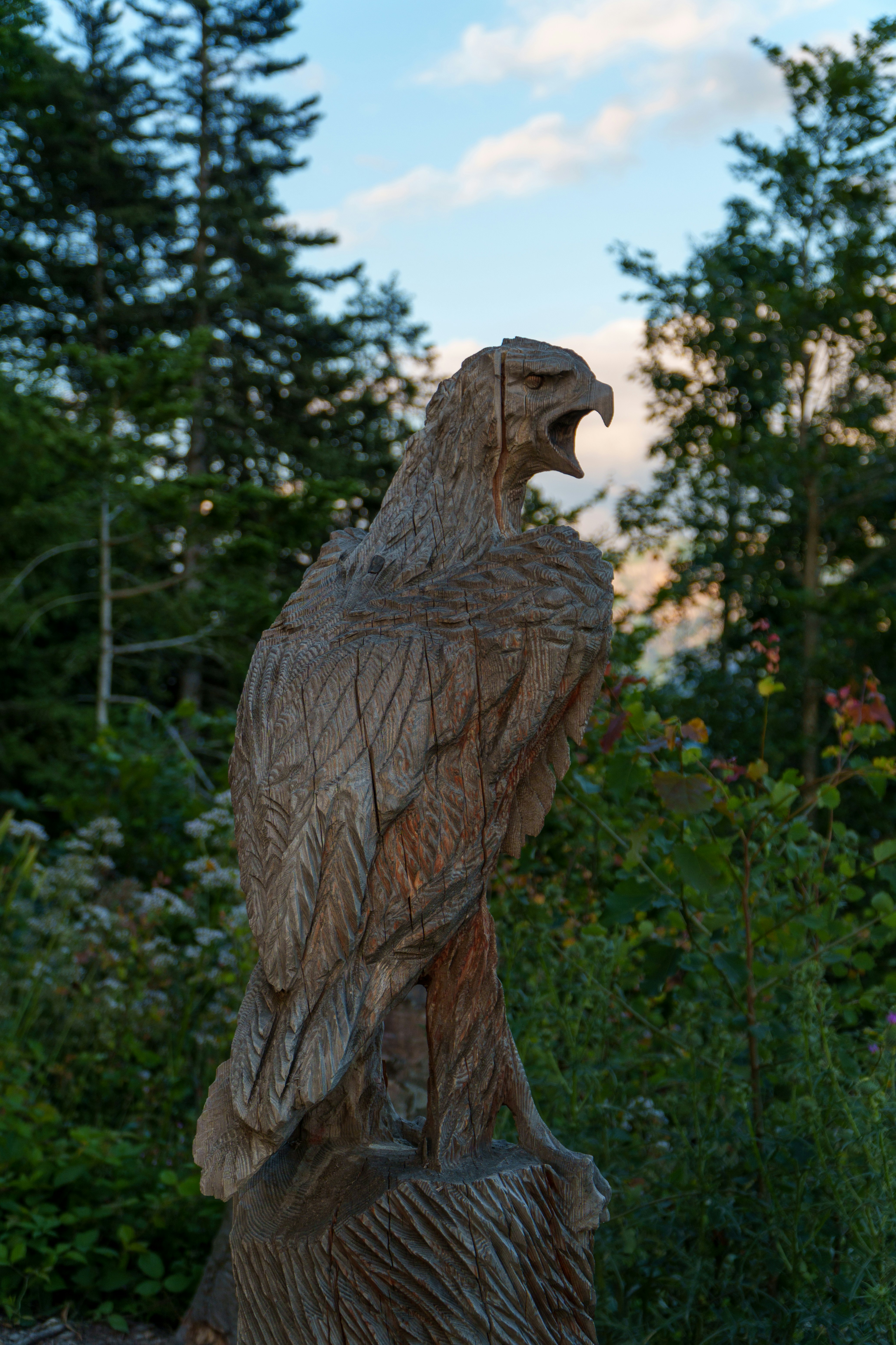 Whittled Sentinel of the Pines: a weathered eagle carving rises among trees at dusk