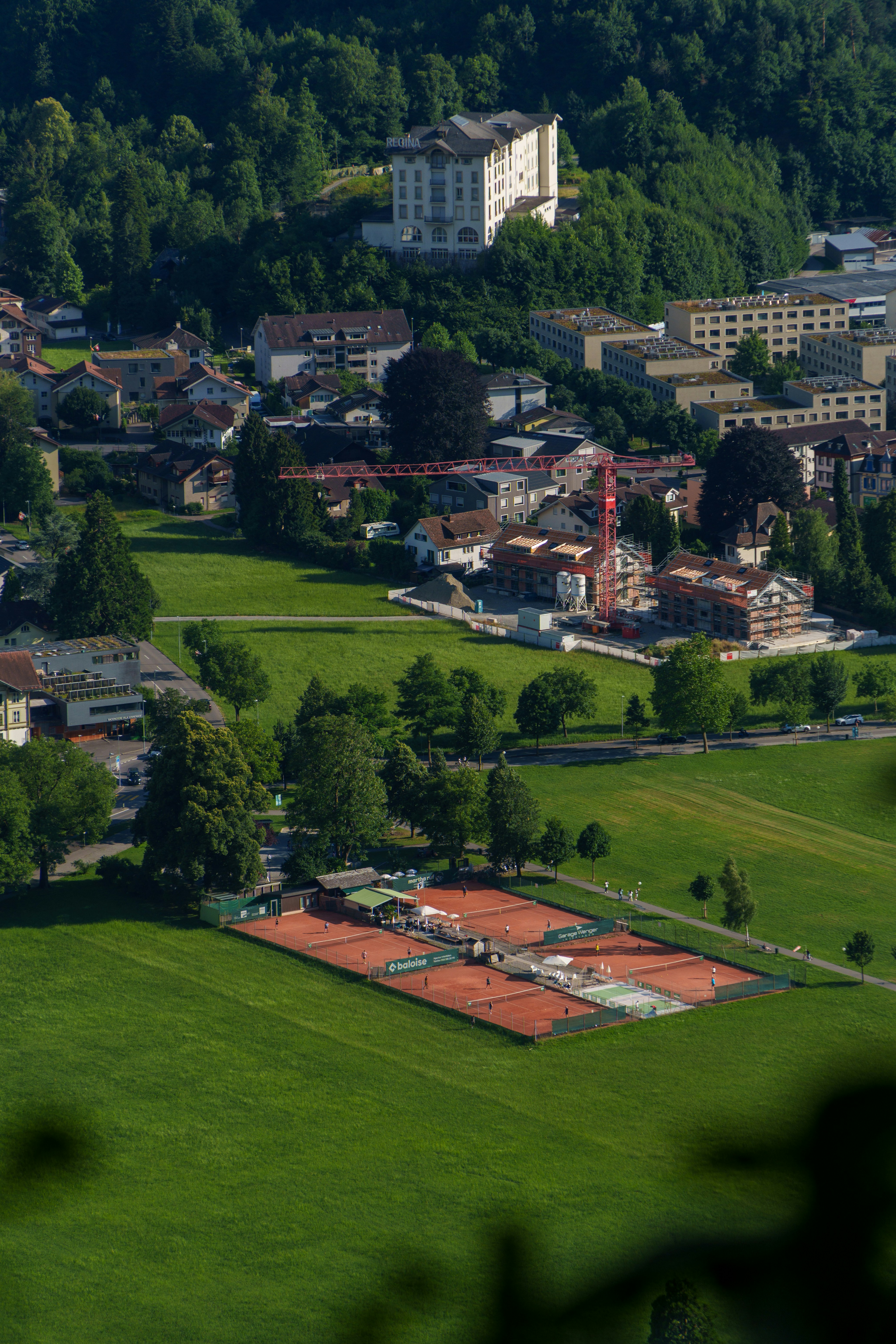 Aerial view of a vibrant tennis court surrounded by lush greenery and urban development, showcasing the blend of sport and nature.