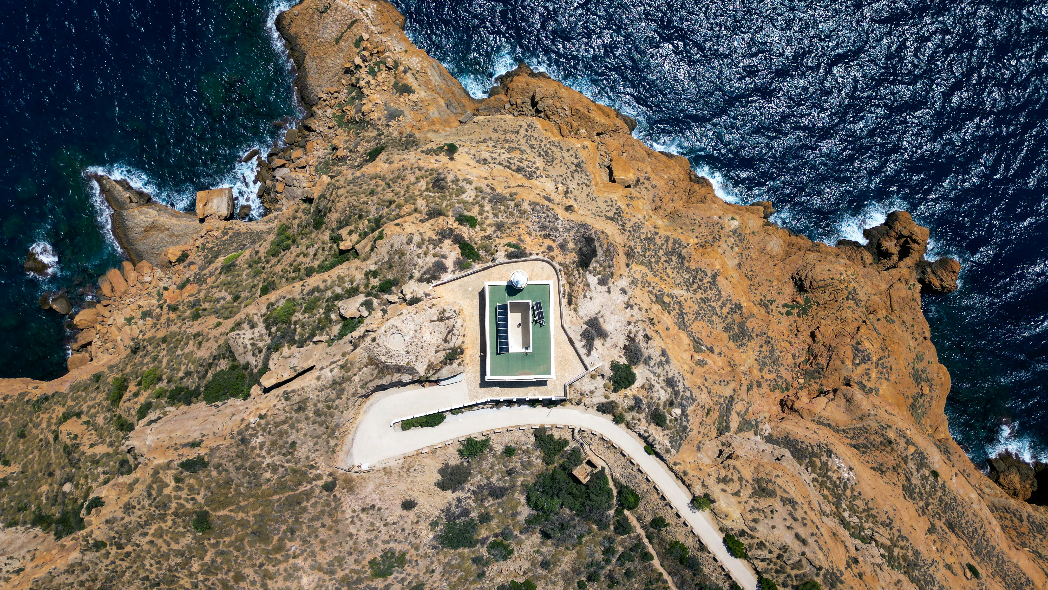 Aerial view of a coastal structure perched on a rugged cliff, surrounded by the deep blue sea and rocky shoreline. The winding path leads to the building, highlighting its isolation.