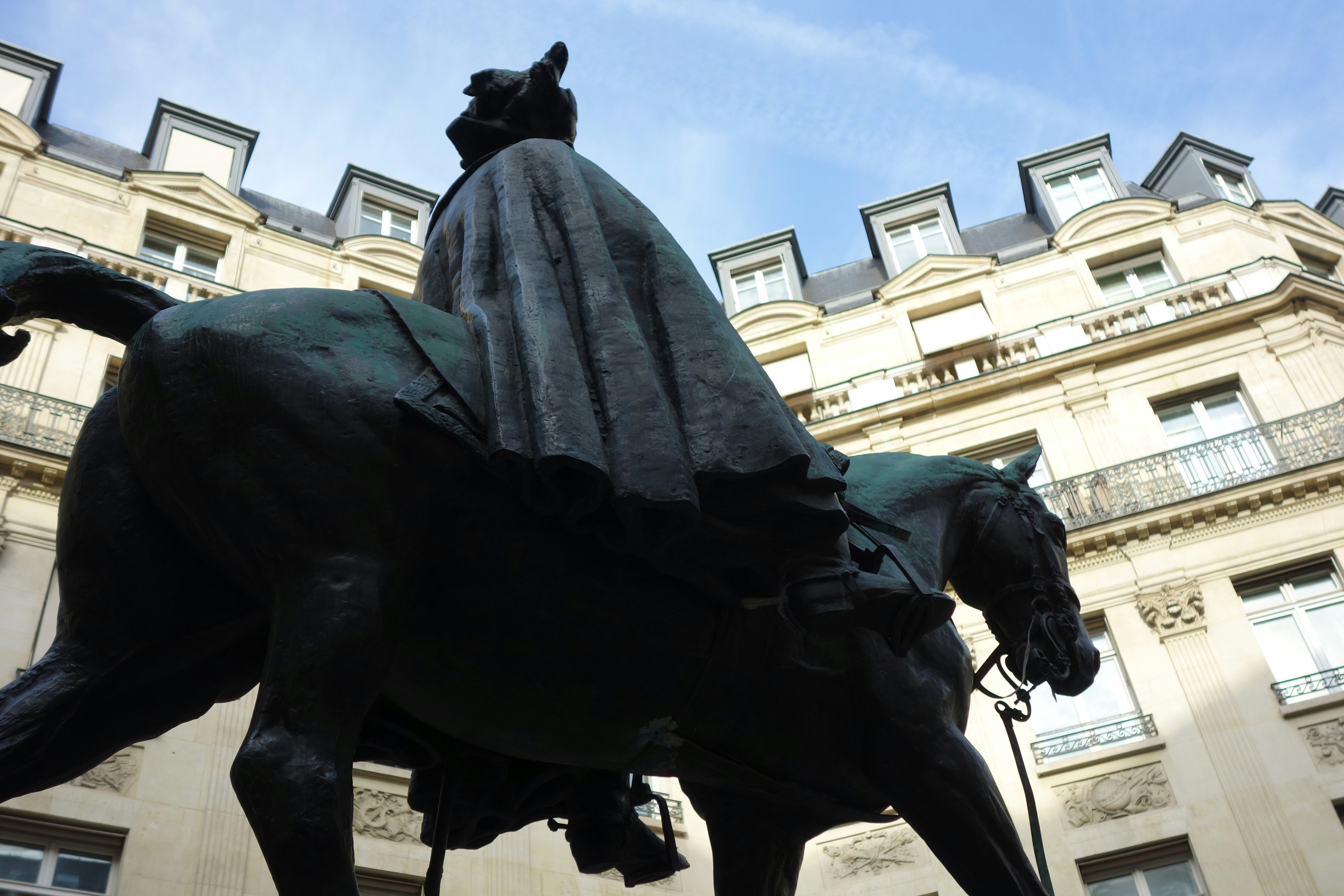Bronze statue of a mounted figure draped in a cloak, set against a backdrop of classic architecture. The perspective emphasizes the grandeur of the sculpture.
