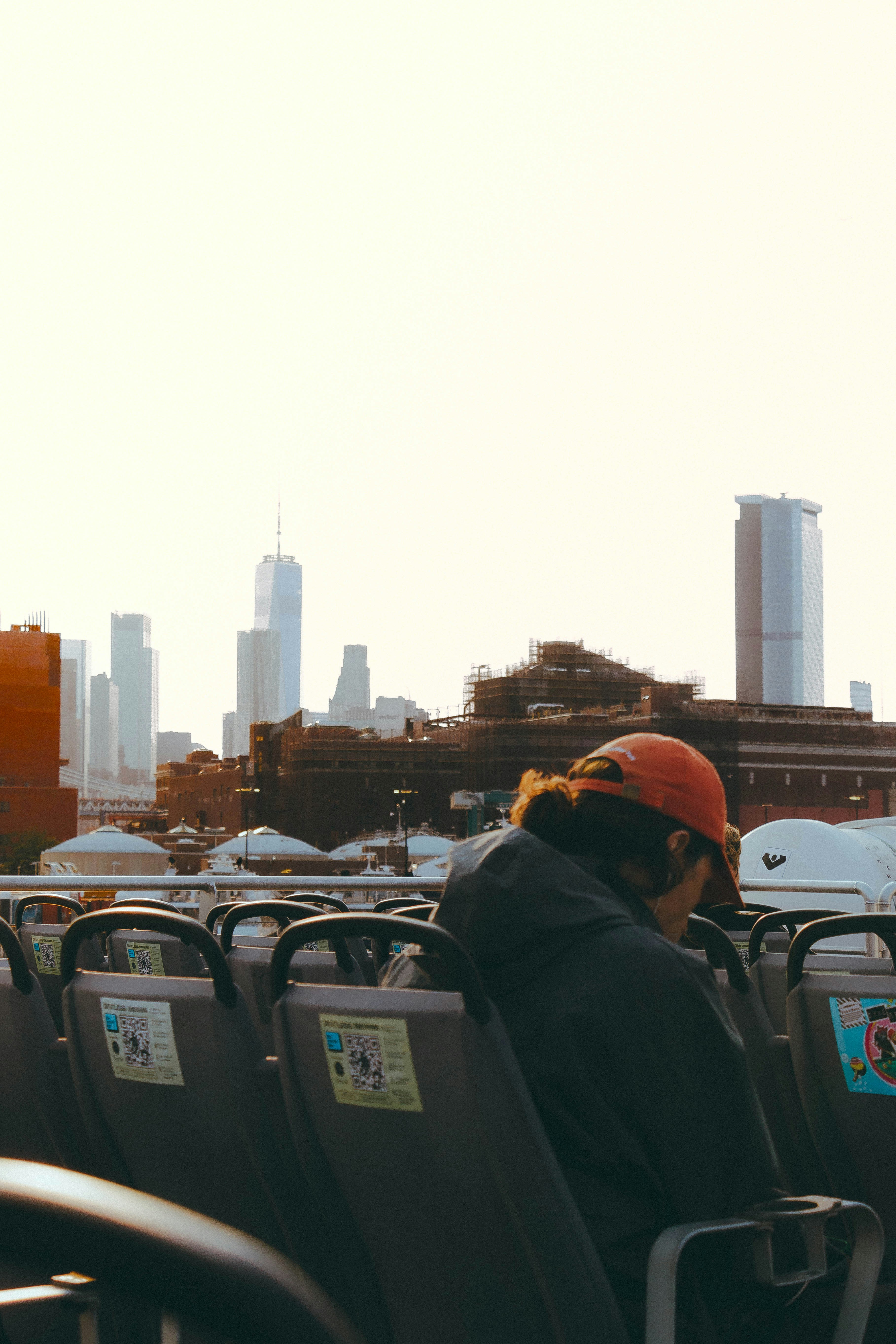 Moody shot of a person on a ferry with New York City skyline in the background at sunset, featuring the One World Trade Center. | A person gazes at a city skyline view.