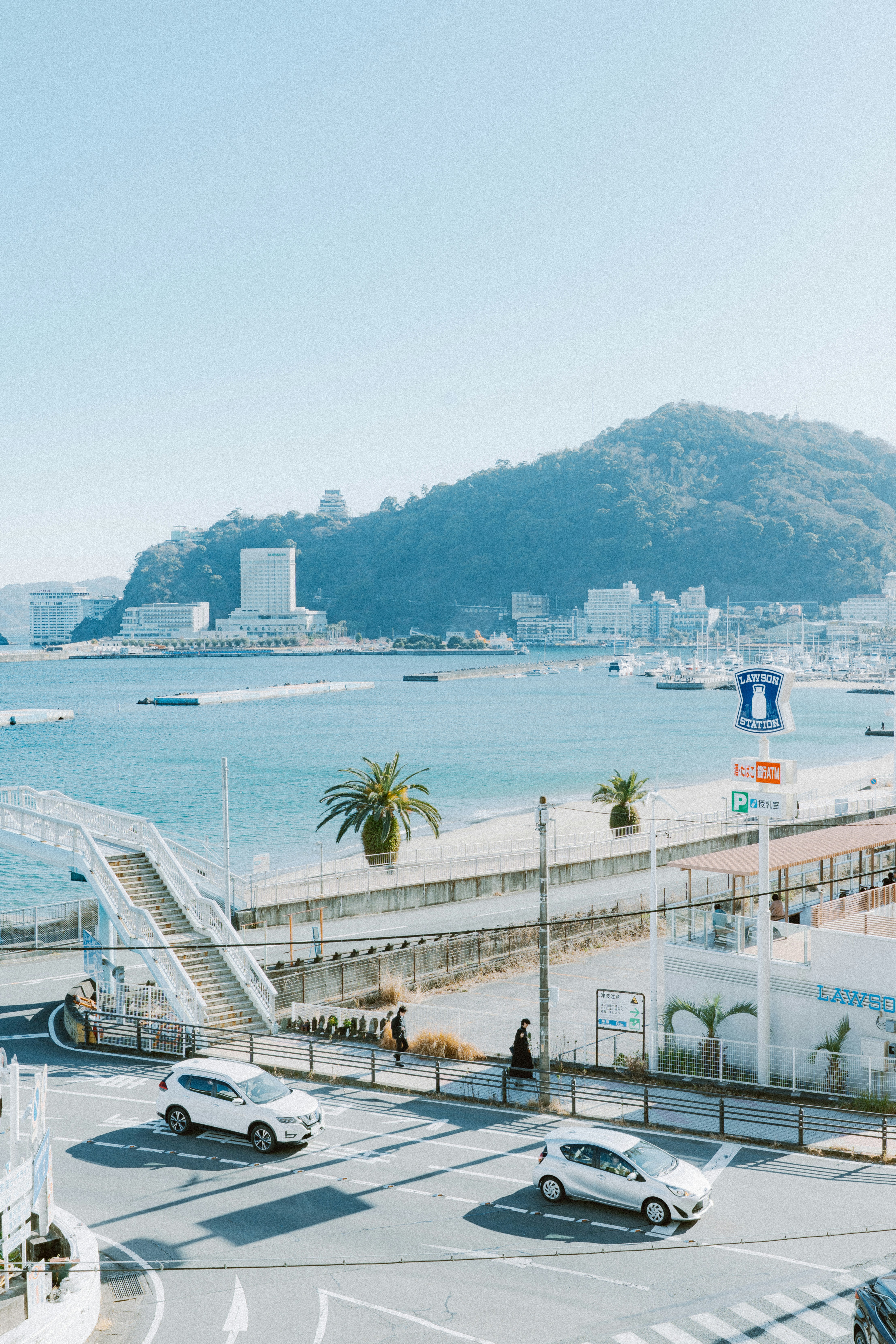 Coastal scene featuring a serene bay with boats, a palm tree, and a backdrop of hills and buildings. The image captures the essence of tranquil seaside living.
