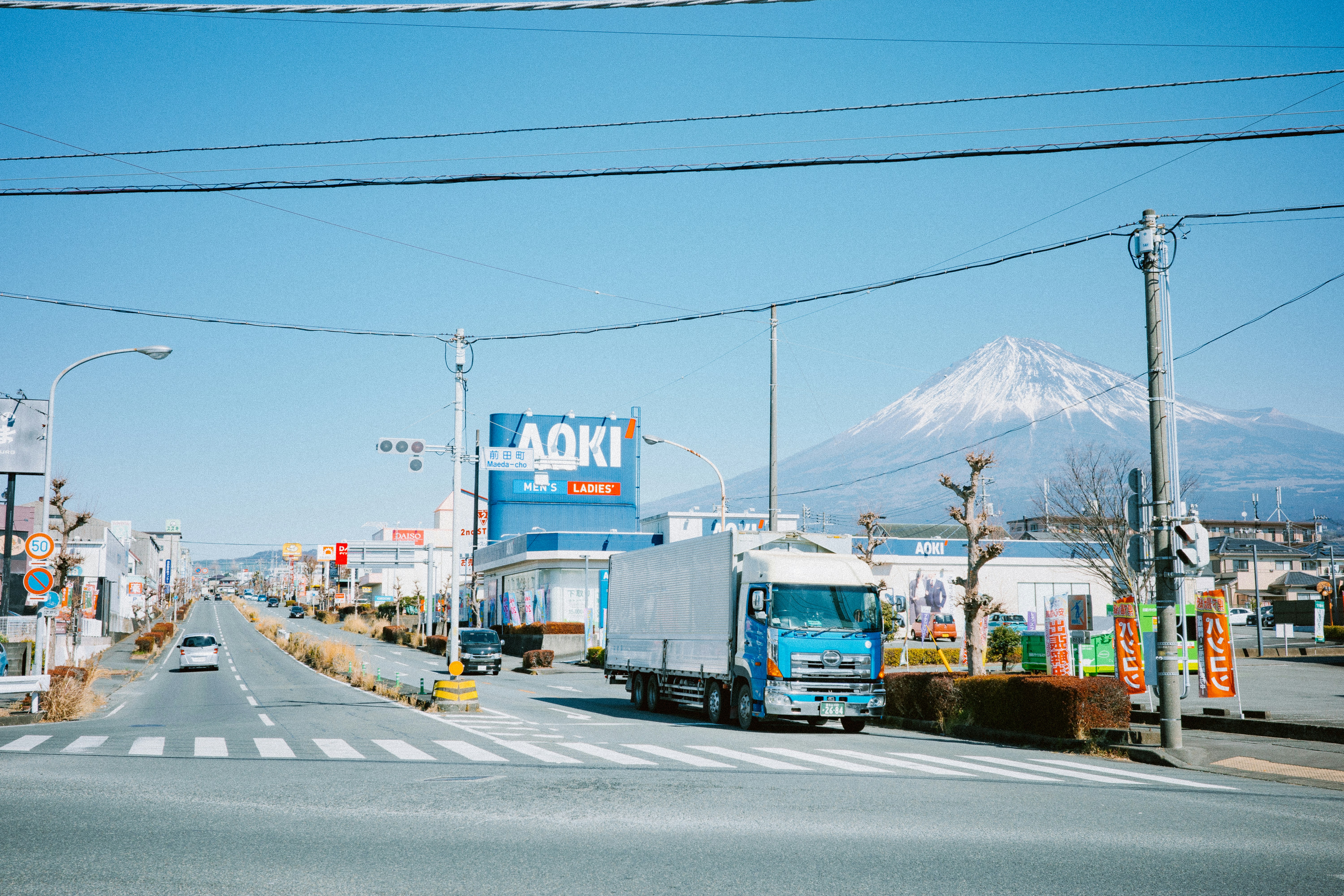 Busy street scene with a truck passing by, framed by commercial signs and the majestic Mount Fuji in the background.