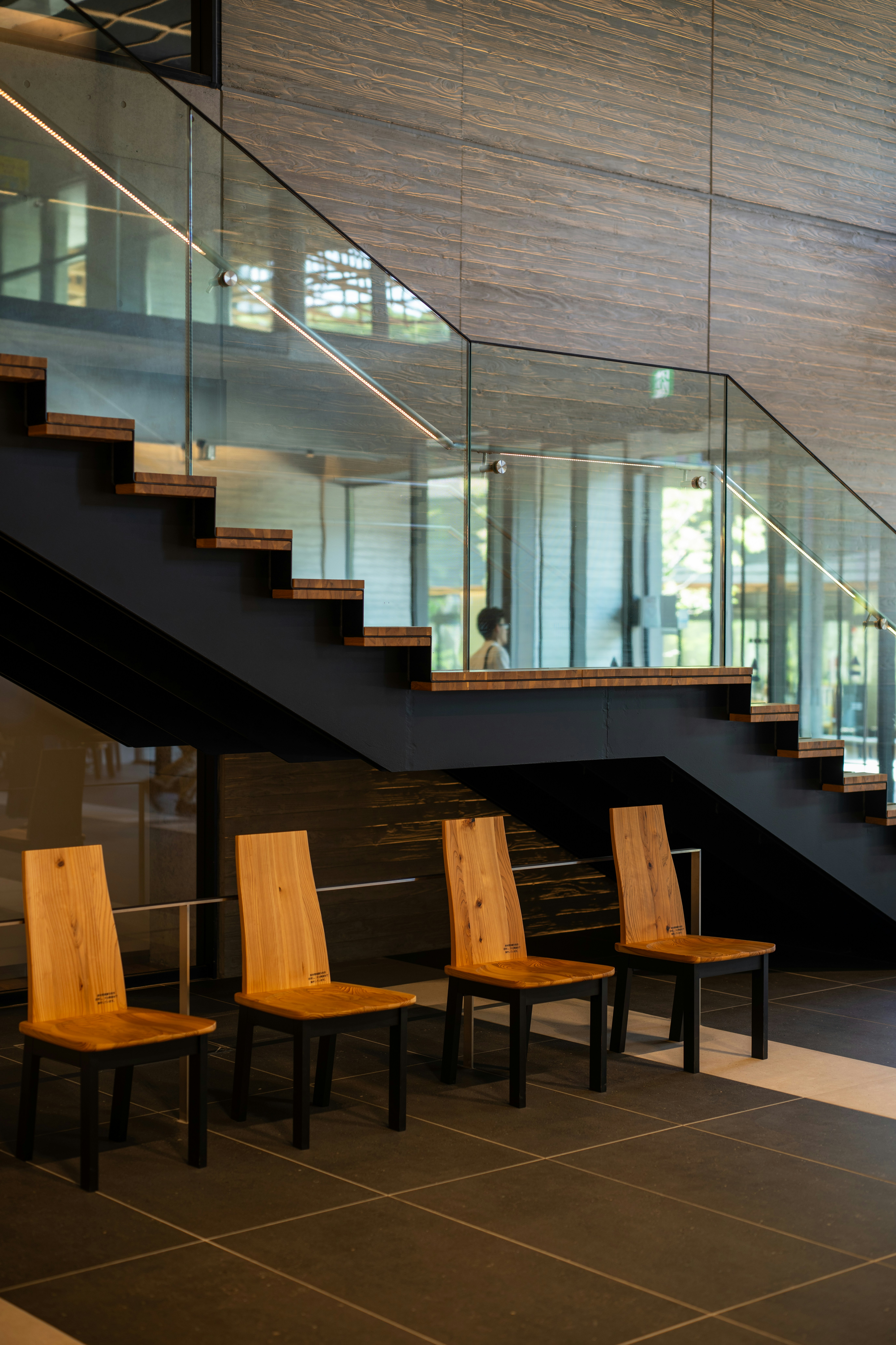 Four wooden chairs arranged neatly on a tiled floor, complemented by a modern staircase and glass railing in a contemporary interior space.