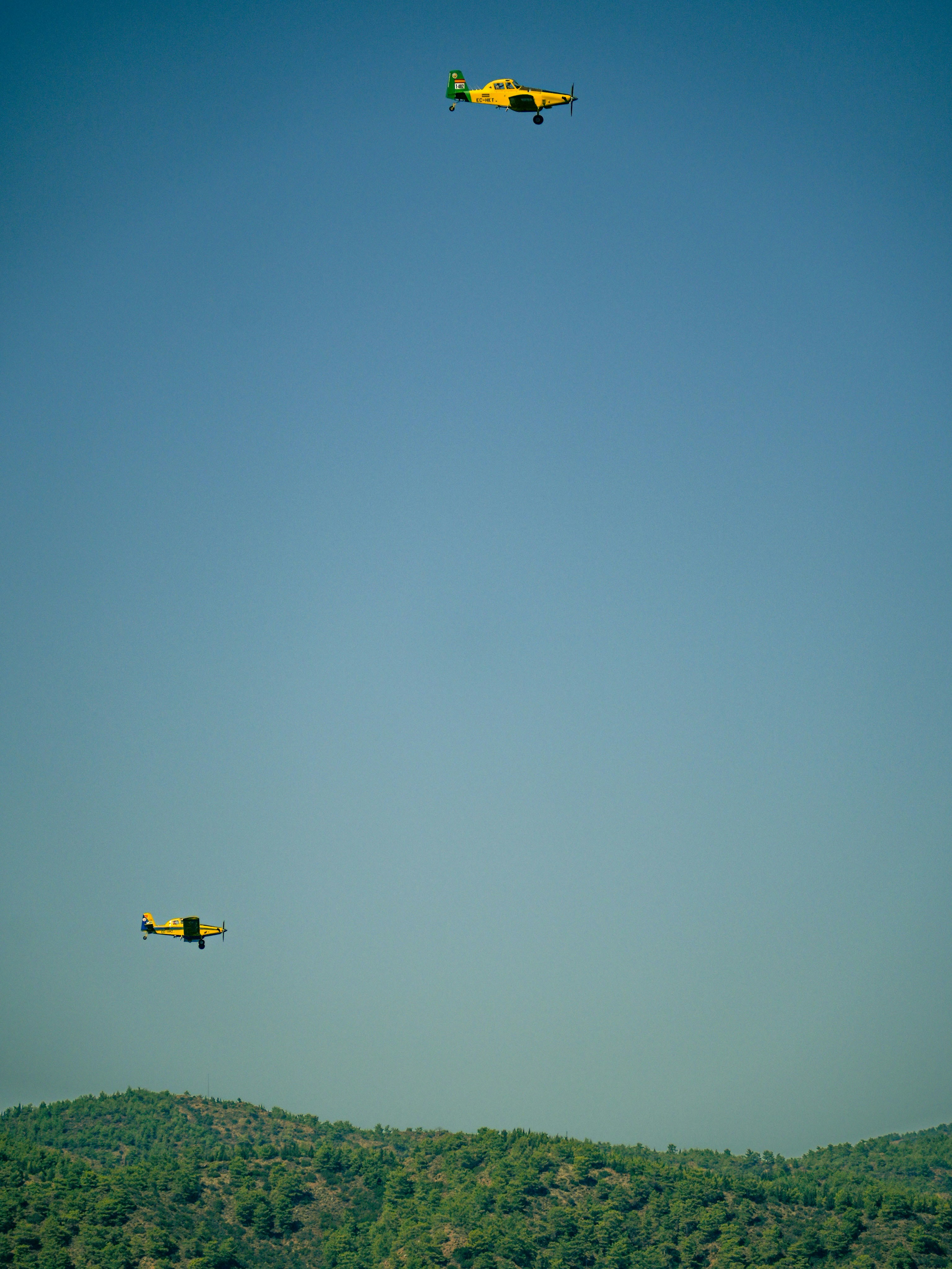 Two yellow light aircraft fly in tandem over a green hill | Two yellow planes fly above a green landscape.