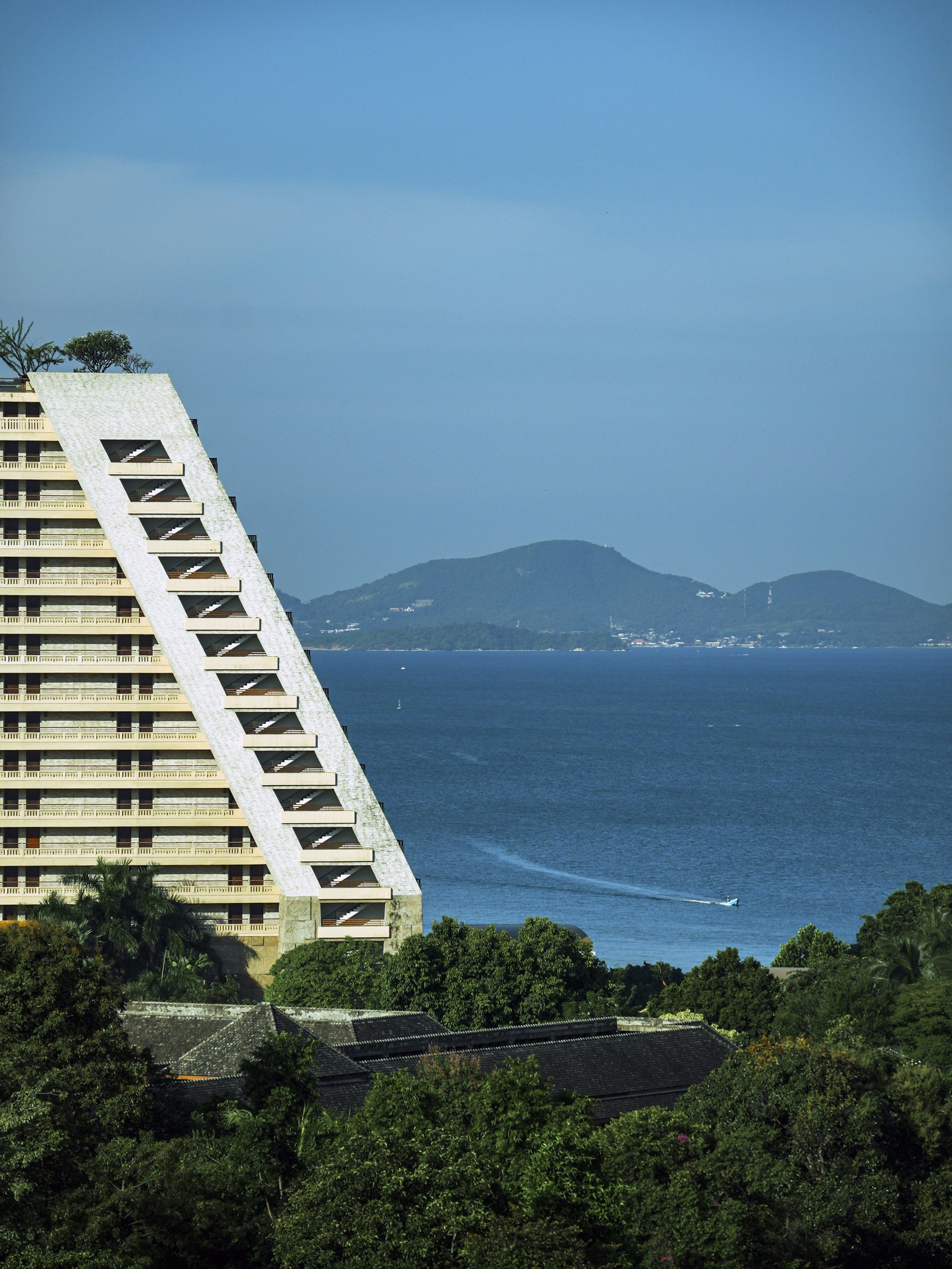 A window with a seaside landscape and a riot of green plants | A white building sits next to the ocean.