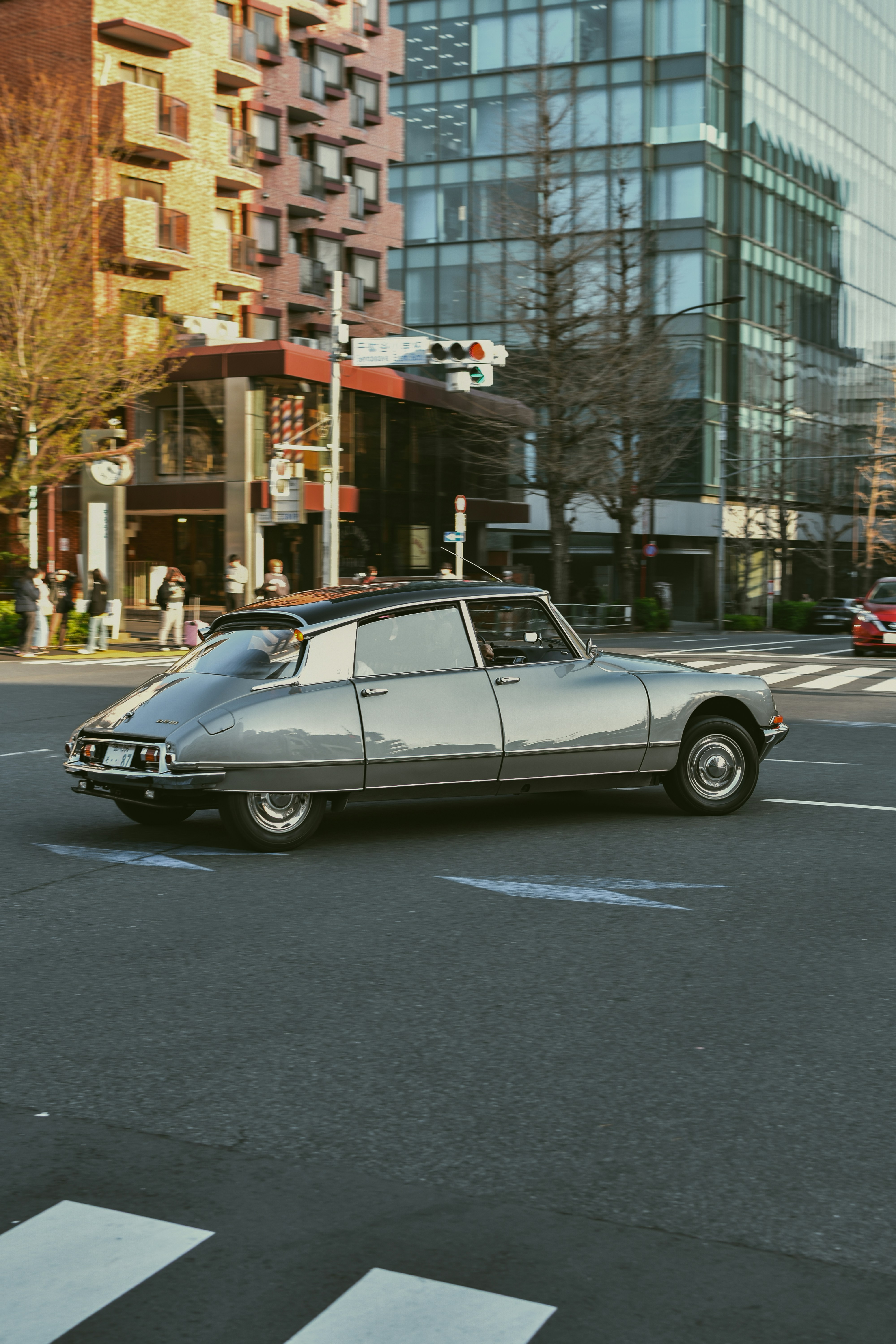 Classic grey car navigating an urban intersection with modern buildings in the background. The scene captures a blend of vintage design and contemporary architecture.