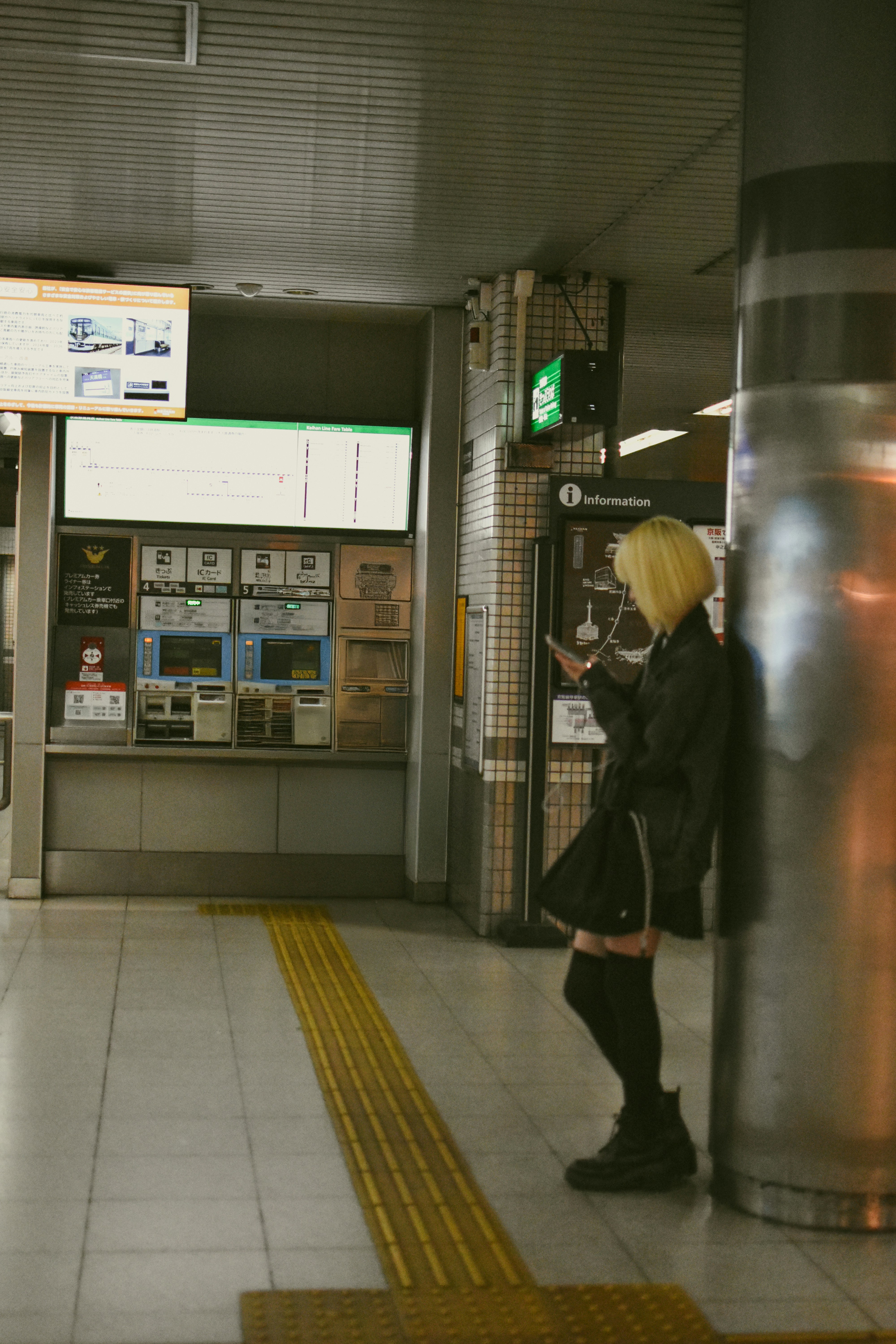 A lone figure with blonde hair stands against a pillar in a subway station, absorbed in their phone while surrounded by ticket machines and digital displays.