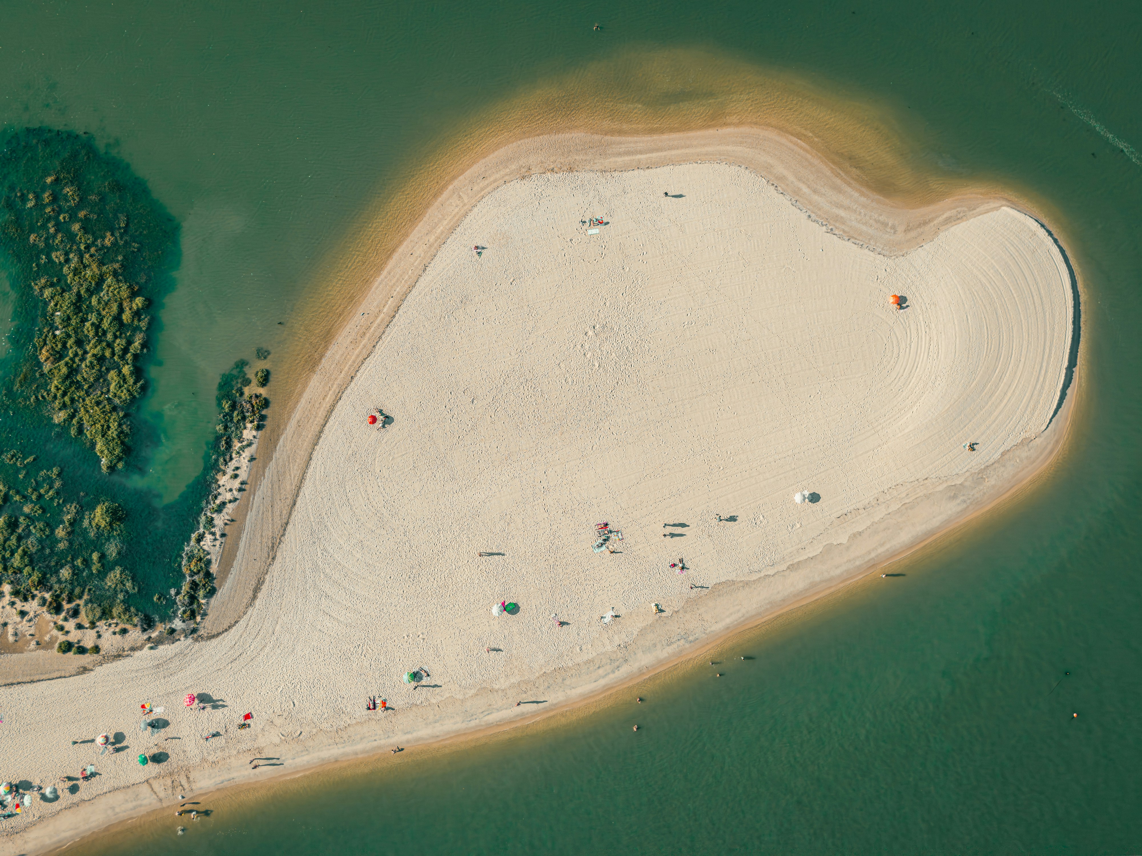 Aerial view of a sandy beach sandbar surrounded by calm waters, with scattered beachgoers enjoying the sun. The lush greenery contrasts with the golden sand.