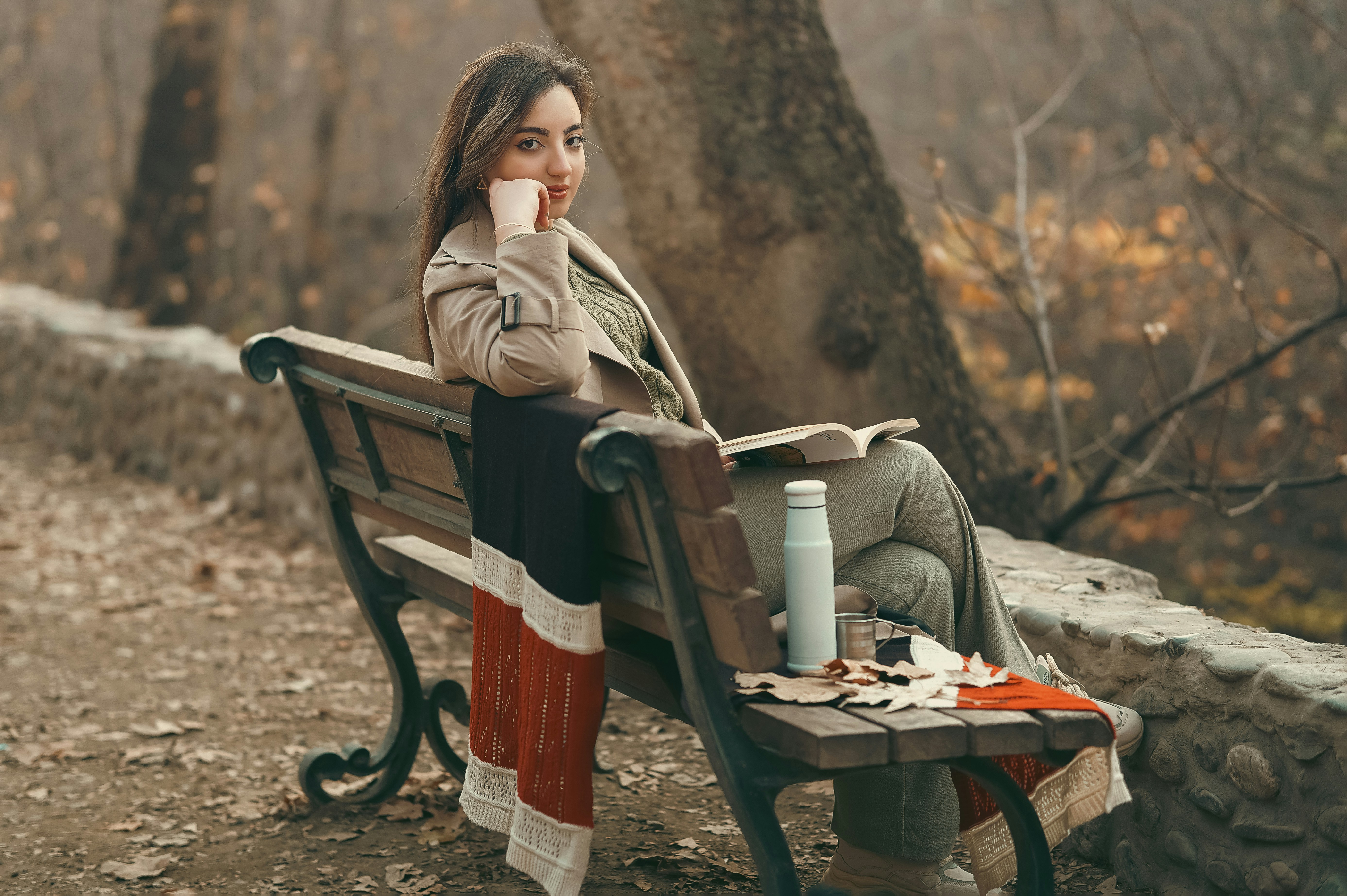 Young woman seated on a park bench, wrapped in a cozy scarf, enjoying a book amidst a tranquil autumn landscape.