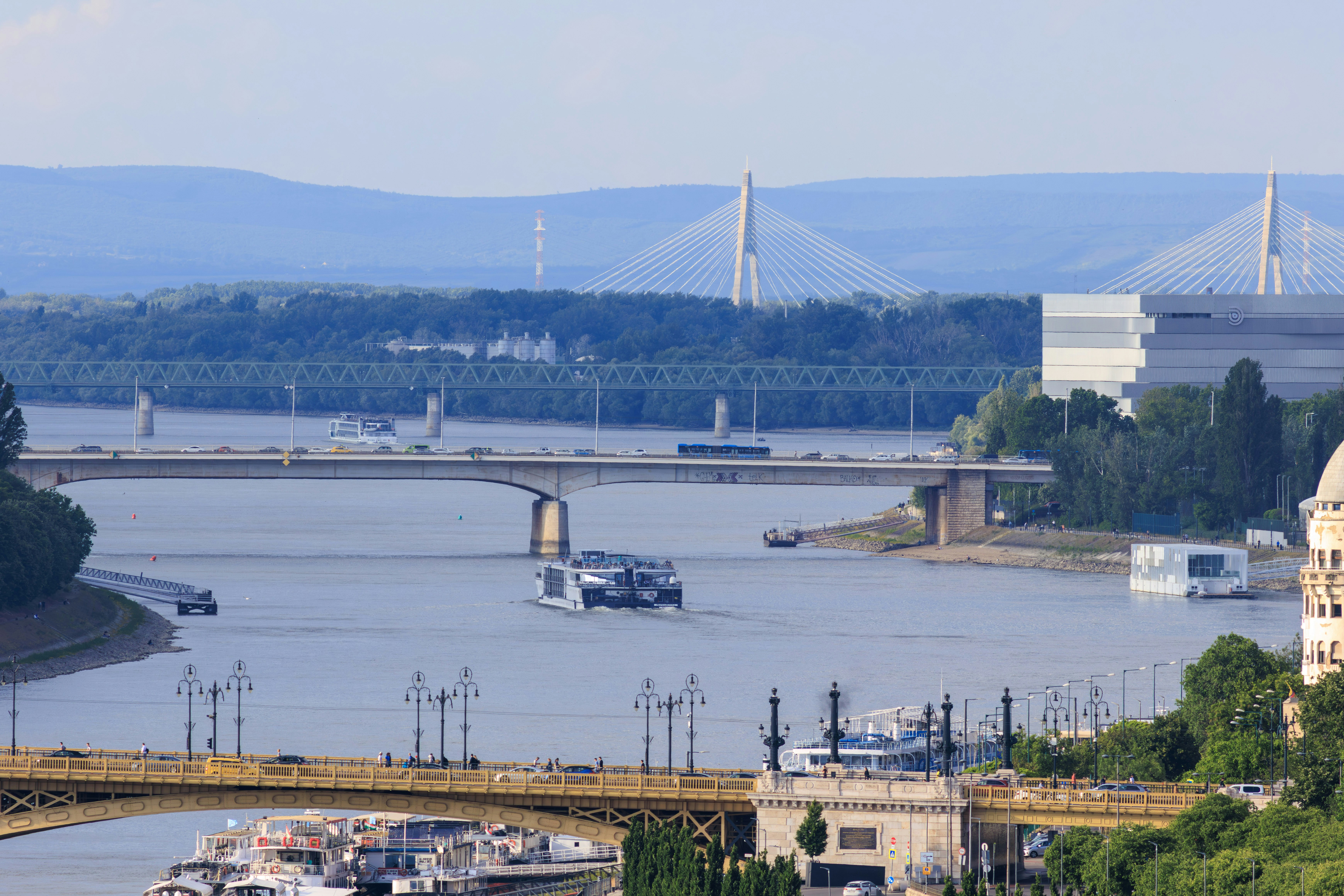 Four bridges from the Buda Castle: Margaret Bridge, Árpád Bridge, Northern Connecting Railway Bridge, Megyeri Bridge | Bridges span a river with a boat sailing on it.