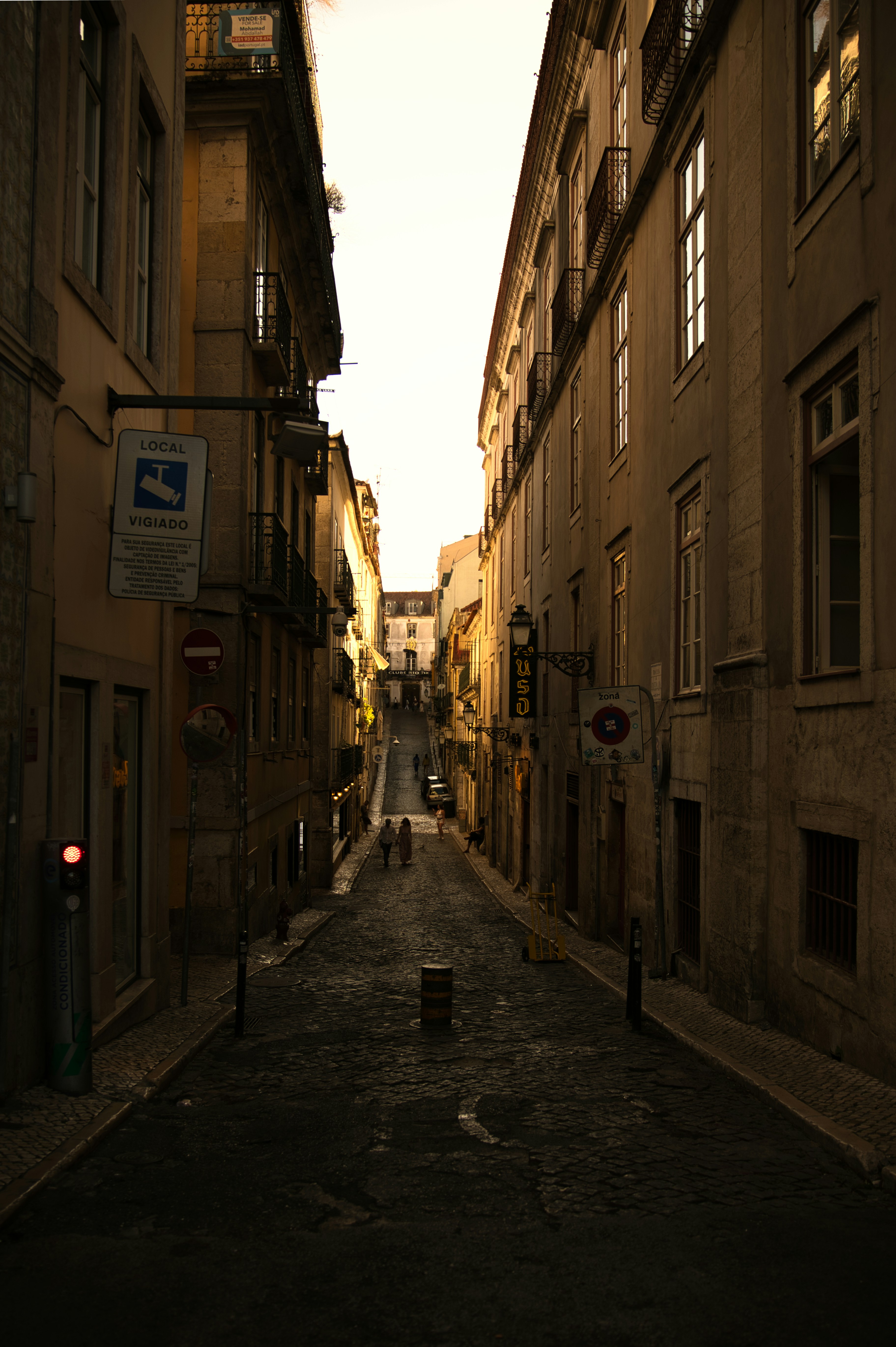 Narrow cobblestone street in Lisbon at dusk, framed by historic buildings and illuminated by soft golden light.