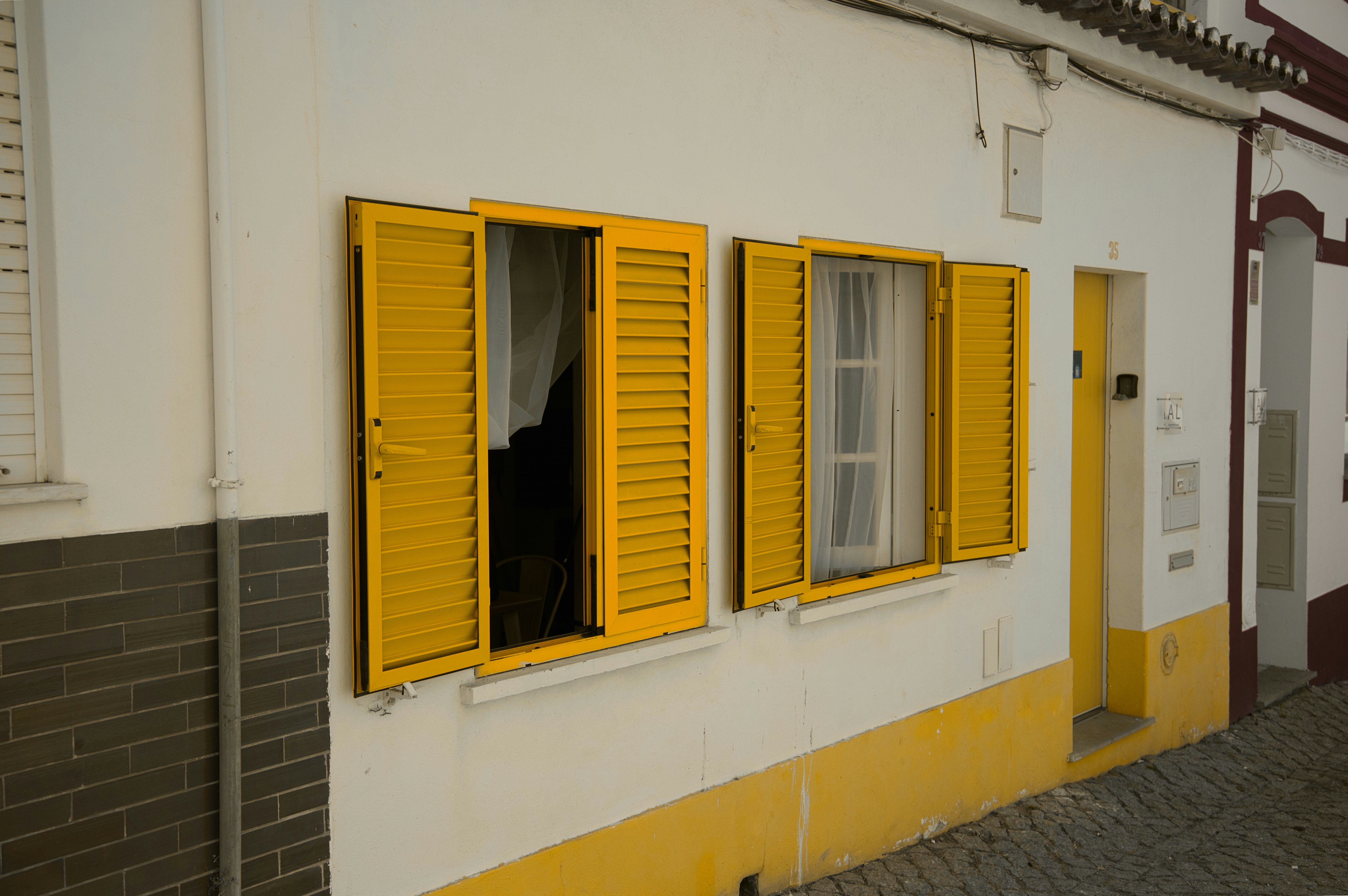 Street Photography in Lagos, Portugal – Urbanes Leben an der Algarve In den malerischen Gassen von Lagos, Portugal, treffe ich auf Szenen, die das wahre Leben erzählen – ungestellt, roh und voller Geschichte. Als leidenschaftlicher Street Photographer zieht mich die Altstadt von Lagos sofort in ihren Bann: verwitterte Fassaden, enge Pflasterstraßen, kleine Cafés, Marktstände und das Spiel von Licht und Schatten zwischen traditioneller portugiesischer Architektur und modernem Stadtleben. Mit meiner Kamera dokumentiere ich das echte urbane Leben an der Algarve – spontane Begegnungen, Straßensz | Yellow shutters adorn a white house exterior.