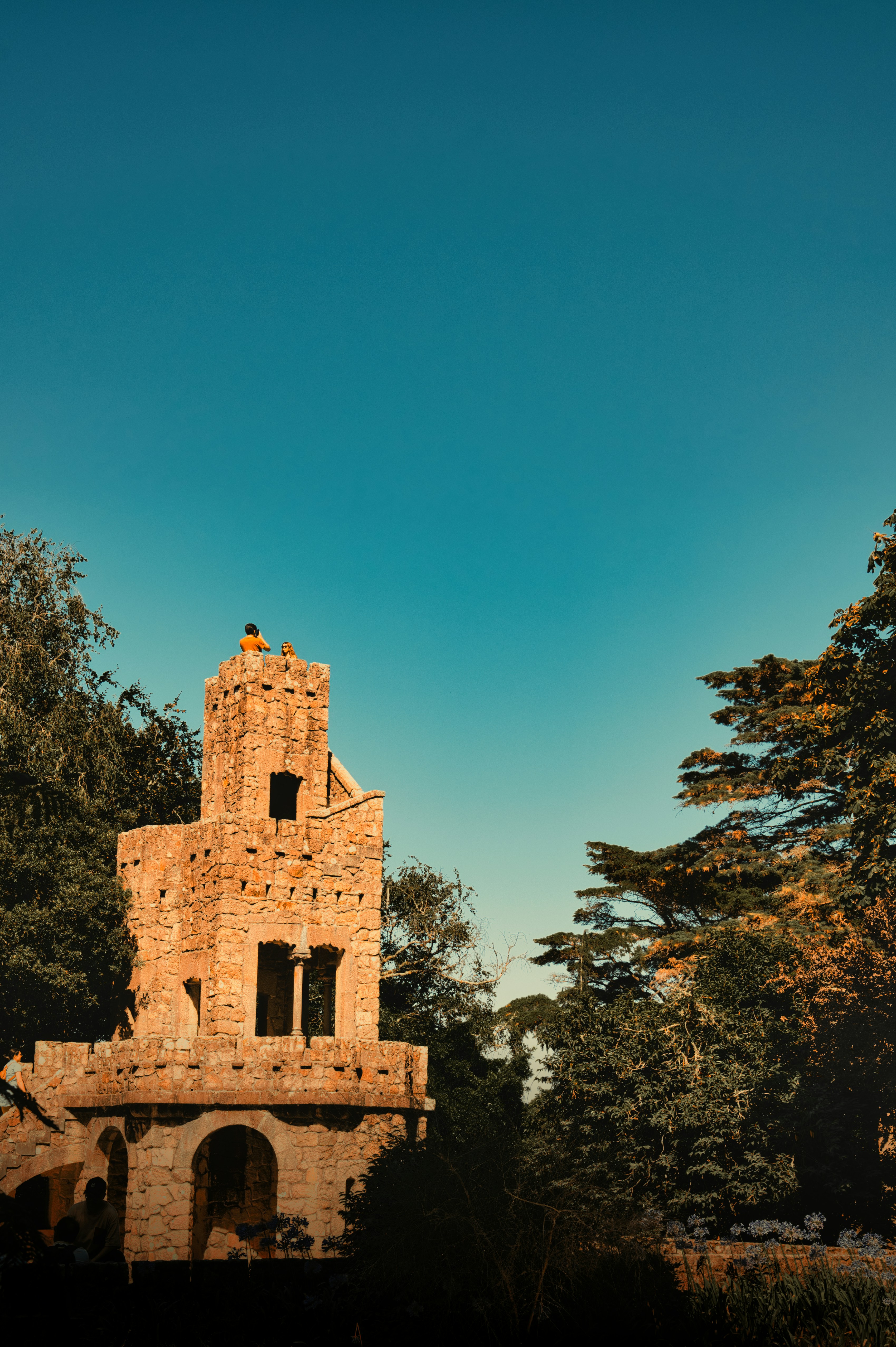 „Quinta da Regaleira, Sintra – fotografiert von Tobias Kaiser mit Nikon Z6 II“ Die geheimnisvolle Quinta da Regaleira nahe dem historischen Zentrum von Sintra, Portugal, entfaltete sich vor meiner Kamera in all ihrer majestätischen Schönheit. Kein Drohnenbild – sondern eine Aufnahme mit Licht und Gefühl, auf einer großzügigen Fläche von vier Hektar. Der neomanuelinische Palast mit gotisch-romanischem Stil, entworfen von Luigi Manini, wirkt wie aus einem romantischen Kunsttraum. Umgeben von tiefen Seen, versteckten Grotten, einem mystischen Initiationsbrunnen und unterirdischen Tunnelsystemen | A stone tower stands tall among lush green trees.