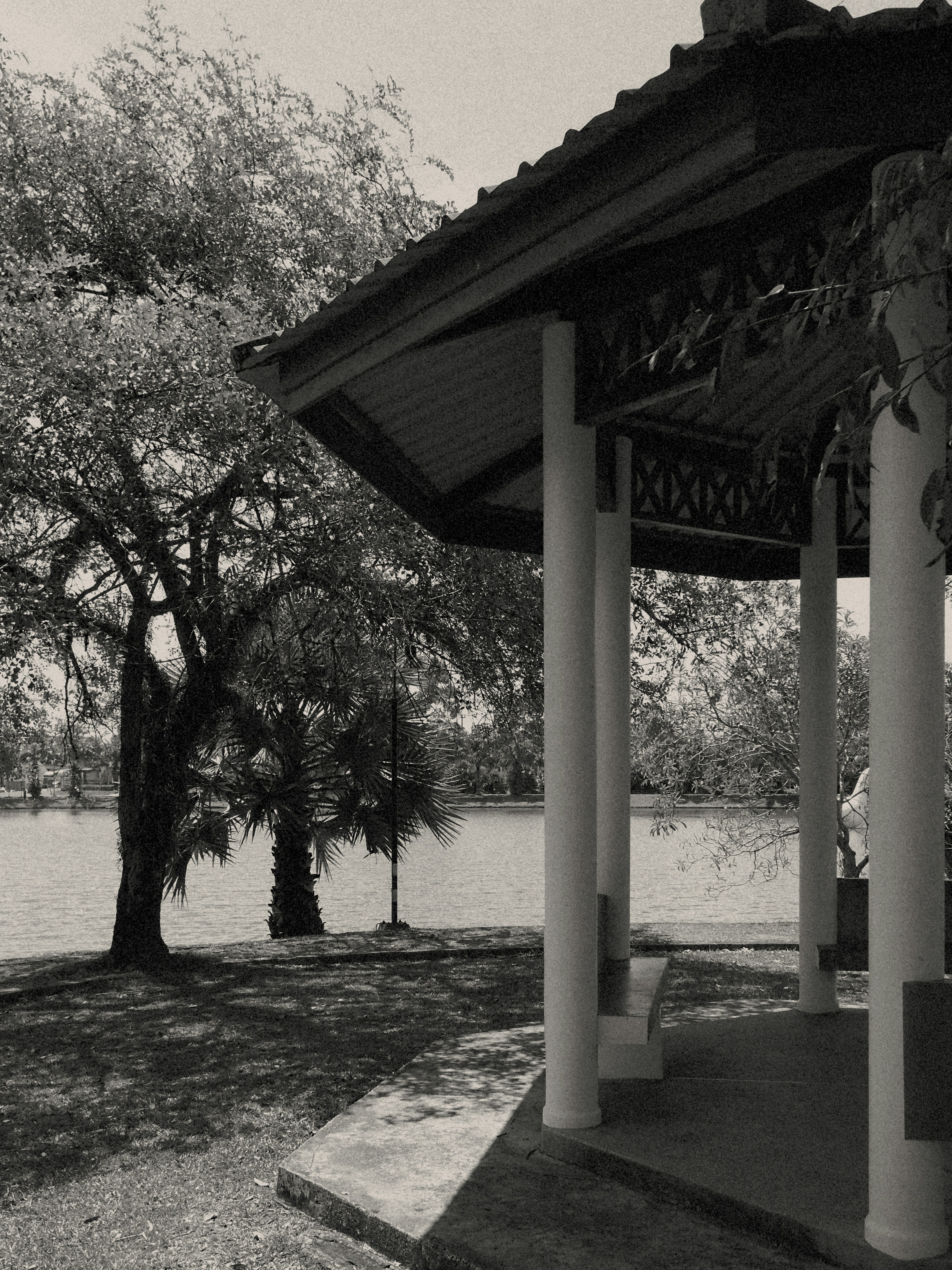 A peaceful riverside pavilion captured in black and white. The image features a shaded structure with classical columns and a slanted roof, framed by trees and calm water in the background. The contrast between light and shadow creates a nostalgic, timeless atmosphere. | Gazebo by a lake on a sunny day.