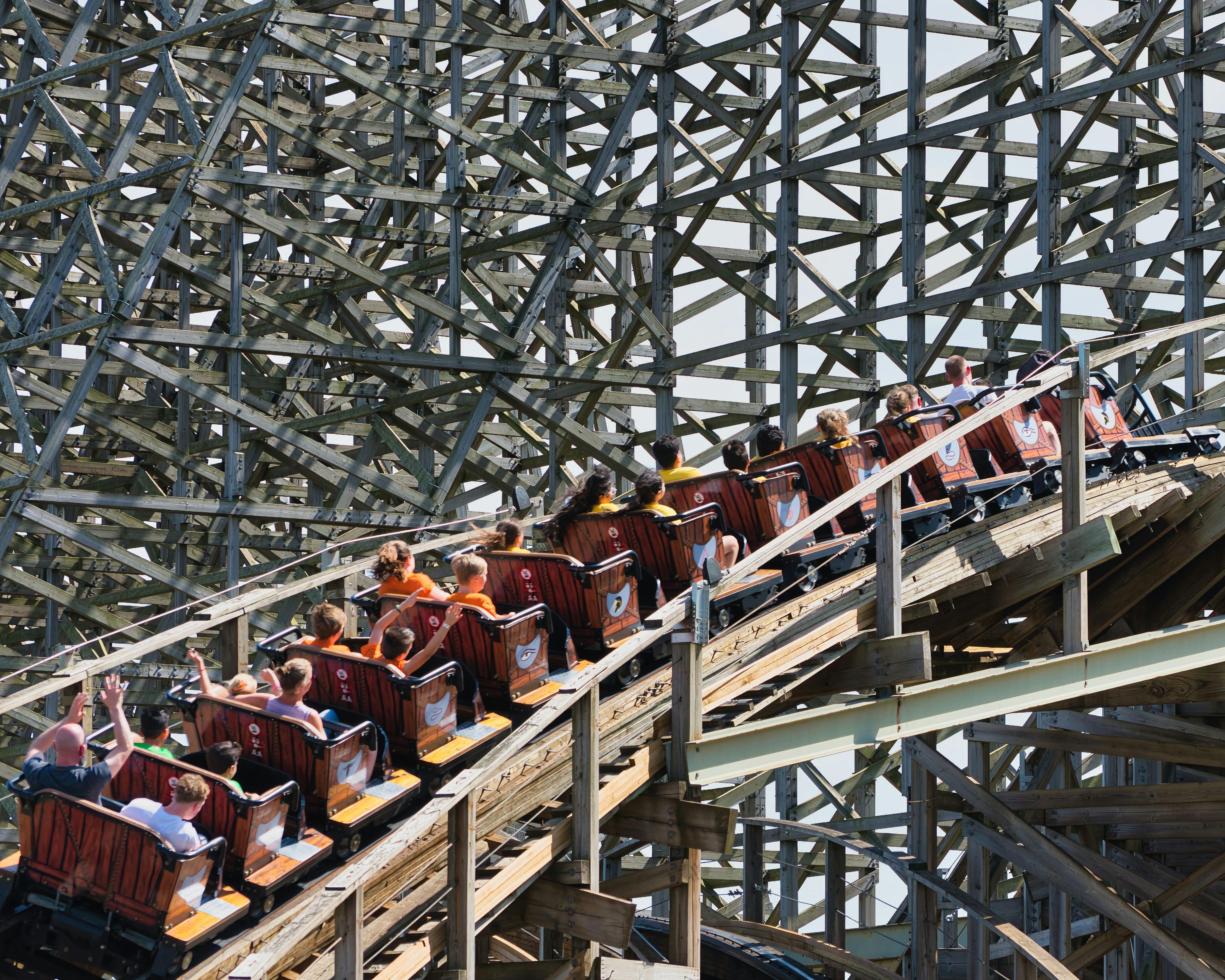 Wooden roller coaster descending with excited riders, showcasing the intricate structure of the track above.